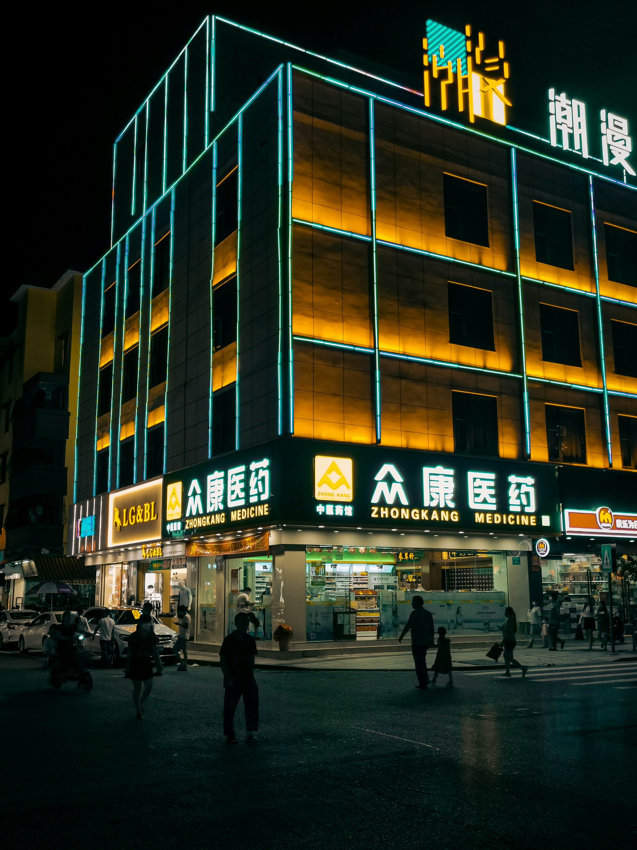 Illuminated storefront of a pharmacy surrounded by bustling pedestrians at night, showcasing vibrant neon lights and urban life.