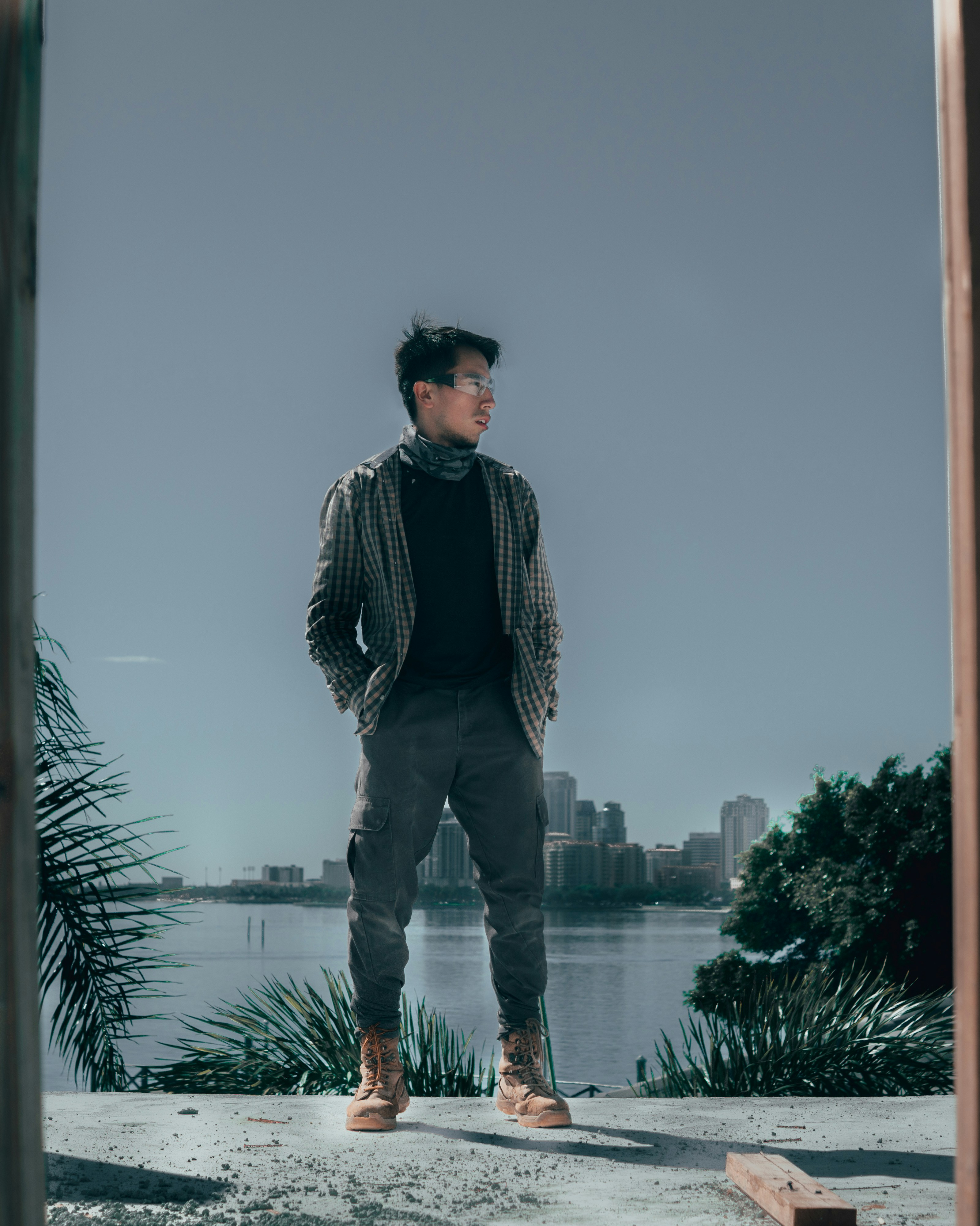 A young man stands confidently on a ledge, framed by lush greenery and a vibrant city skyline in the background. The scene juxtaposes urban life with natural elements.