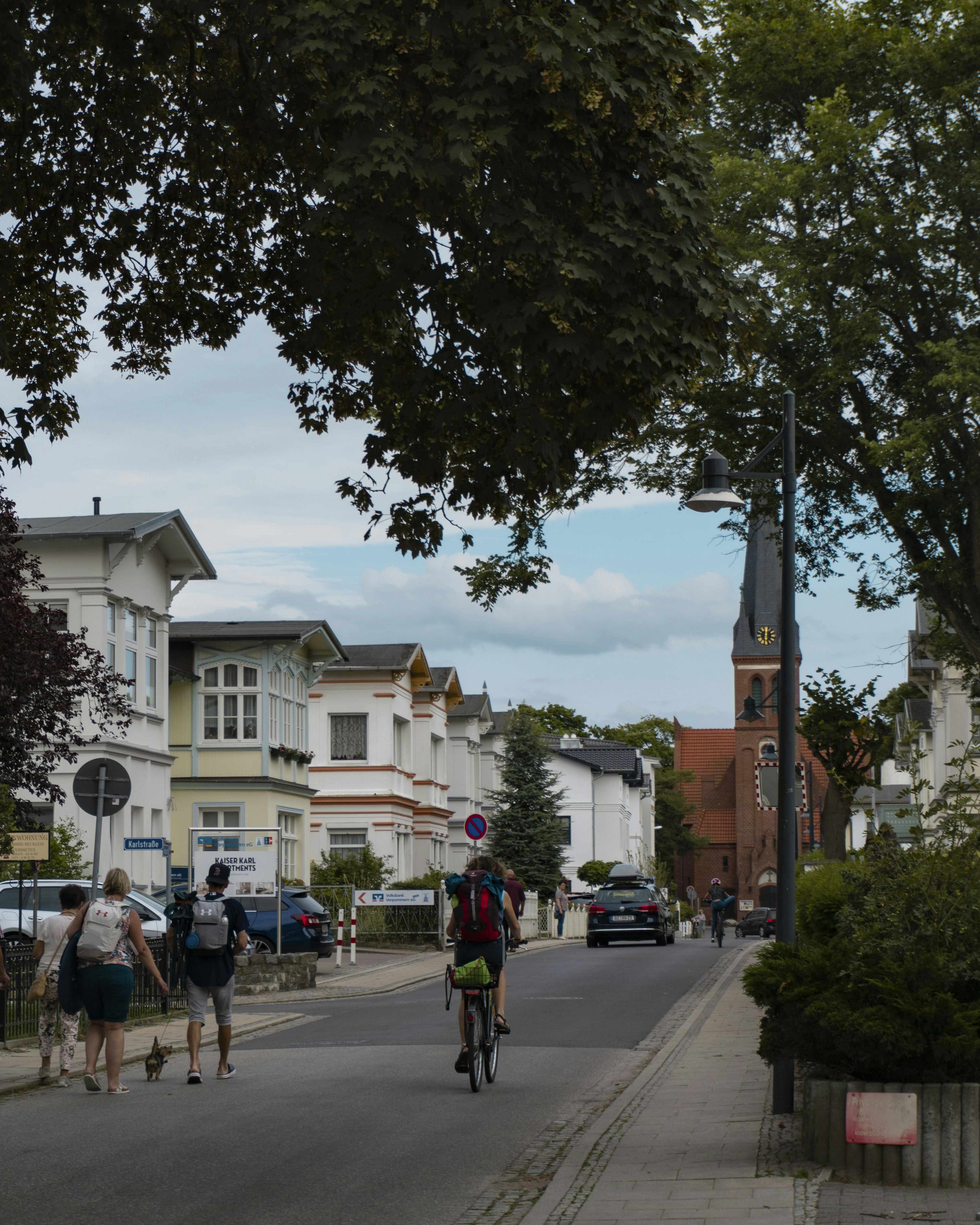 Pedestrians and a cyclist navigate a quiet street lined with charming houses and greenery, with a church steeple visible in the background.