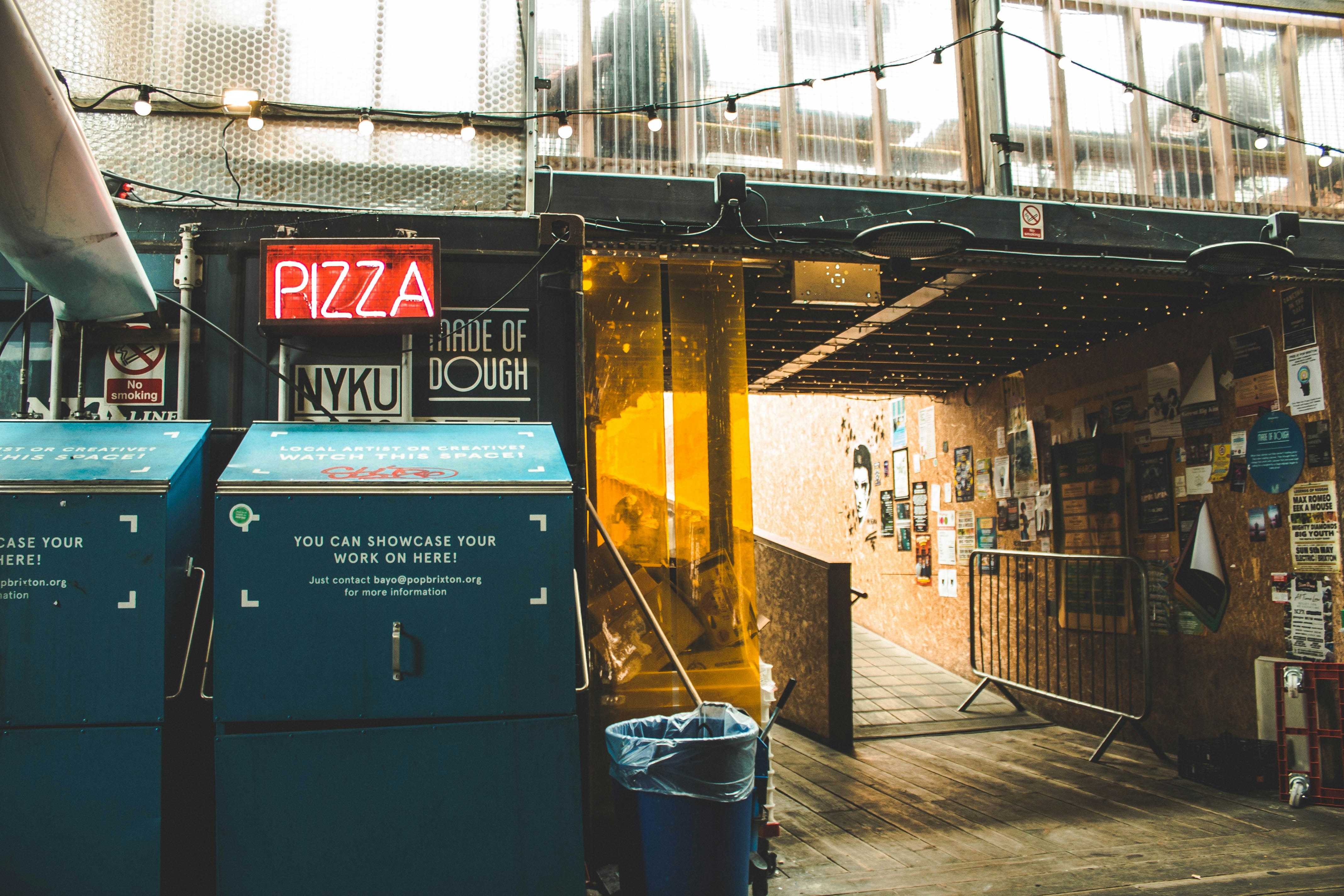 Vibrant food stall area featuring a neon pizza sign, colorful bins, and a warm-lit corridor inviting customers inside.