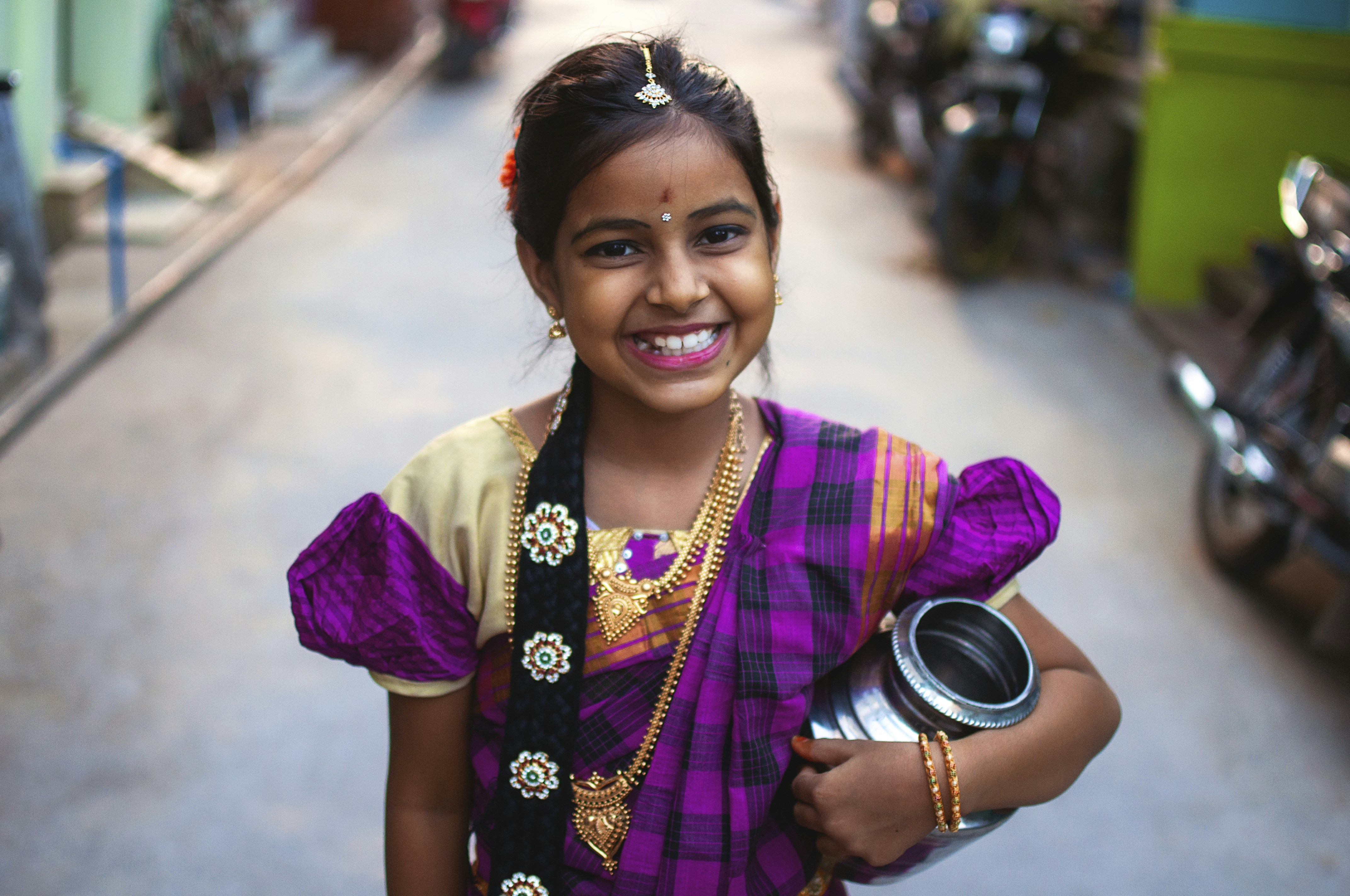 Smiling girl in colorful traditional dress holding a metal container on a bustling street.