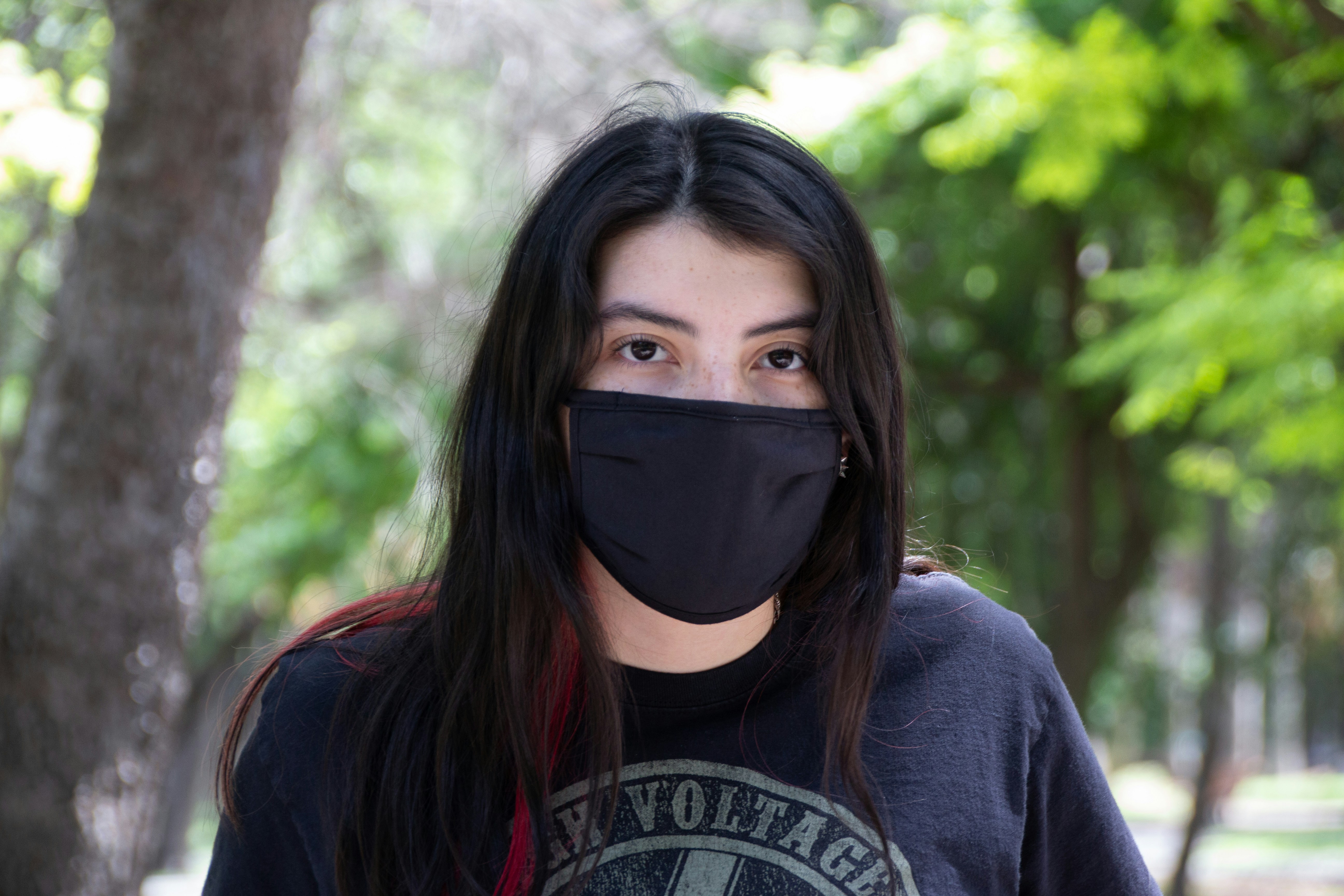 Young person wearing a black mask stands amidst greenery, exuding a calm demeanor. The scene captures a serene moment in a natural setting.