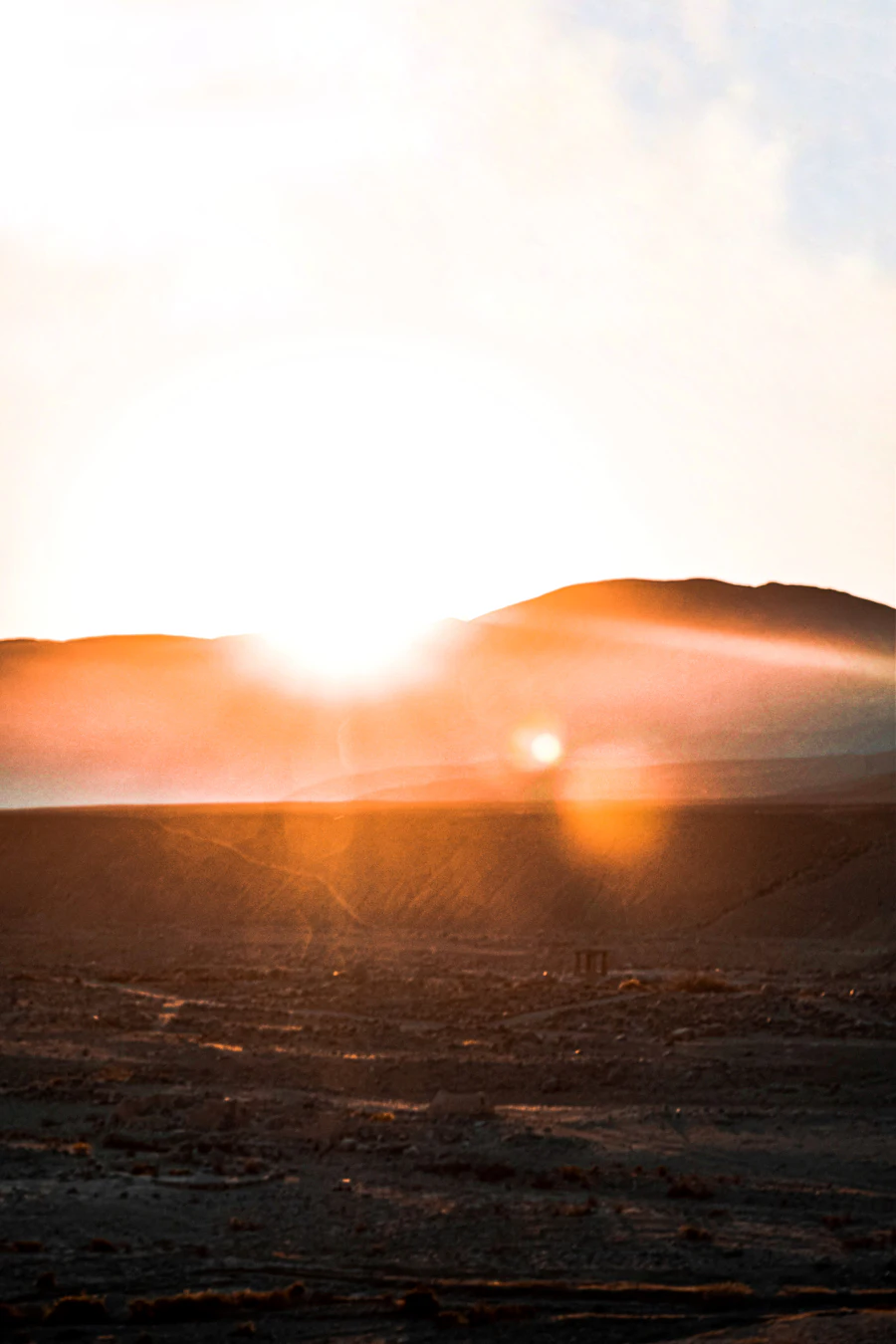 Desert landscape at sunset with the sun perched over a dune, creating warm lens flares across the horizon. The scene emphasizes vast, wind-sculpted terrain and atmospheric glow.