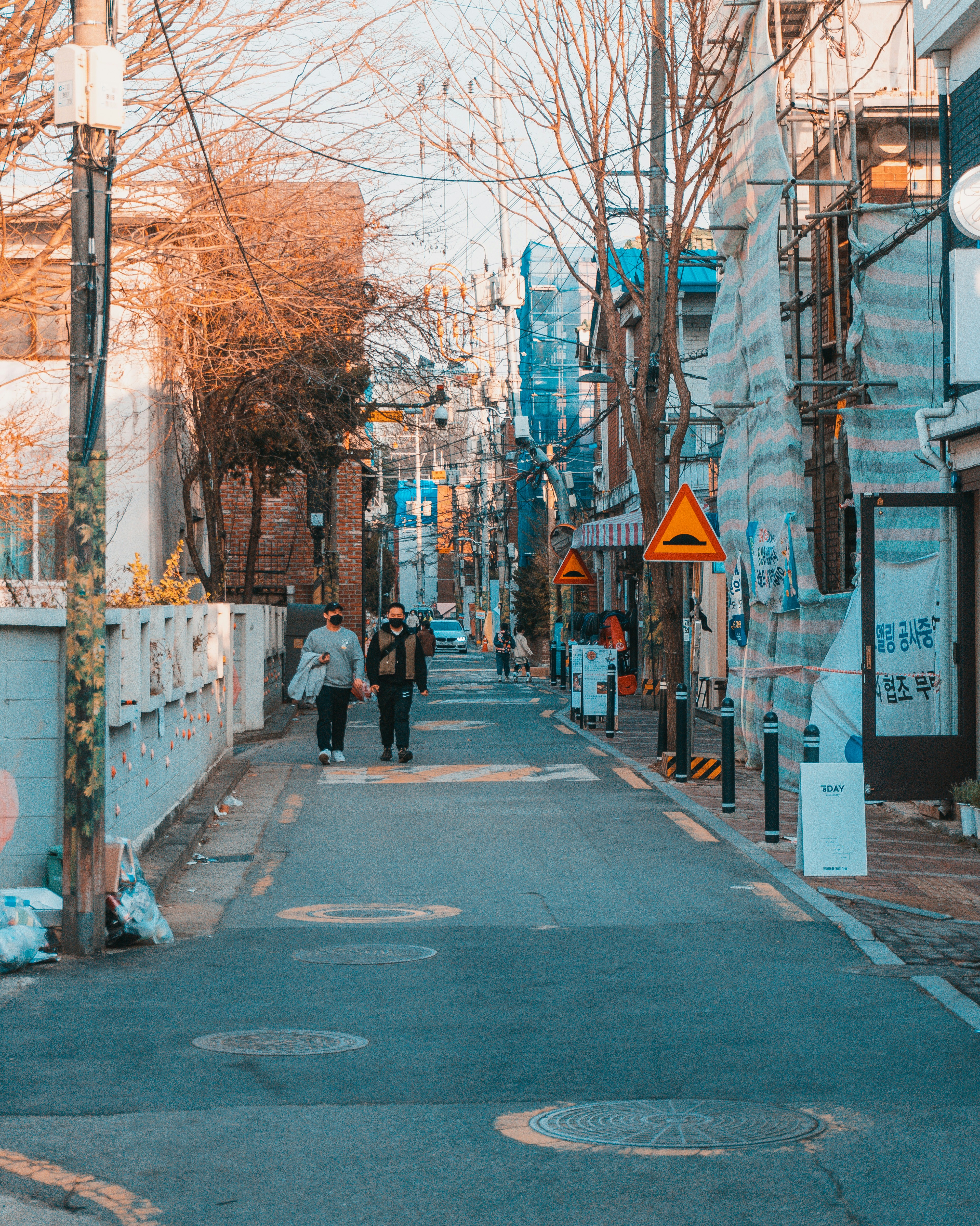 Two pedestrians walking down a tranquil urban alley lined with trees and buildings, showcasing a blend of modern and traditional architecture.
