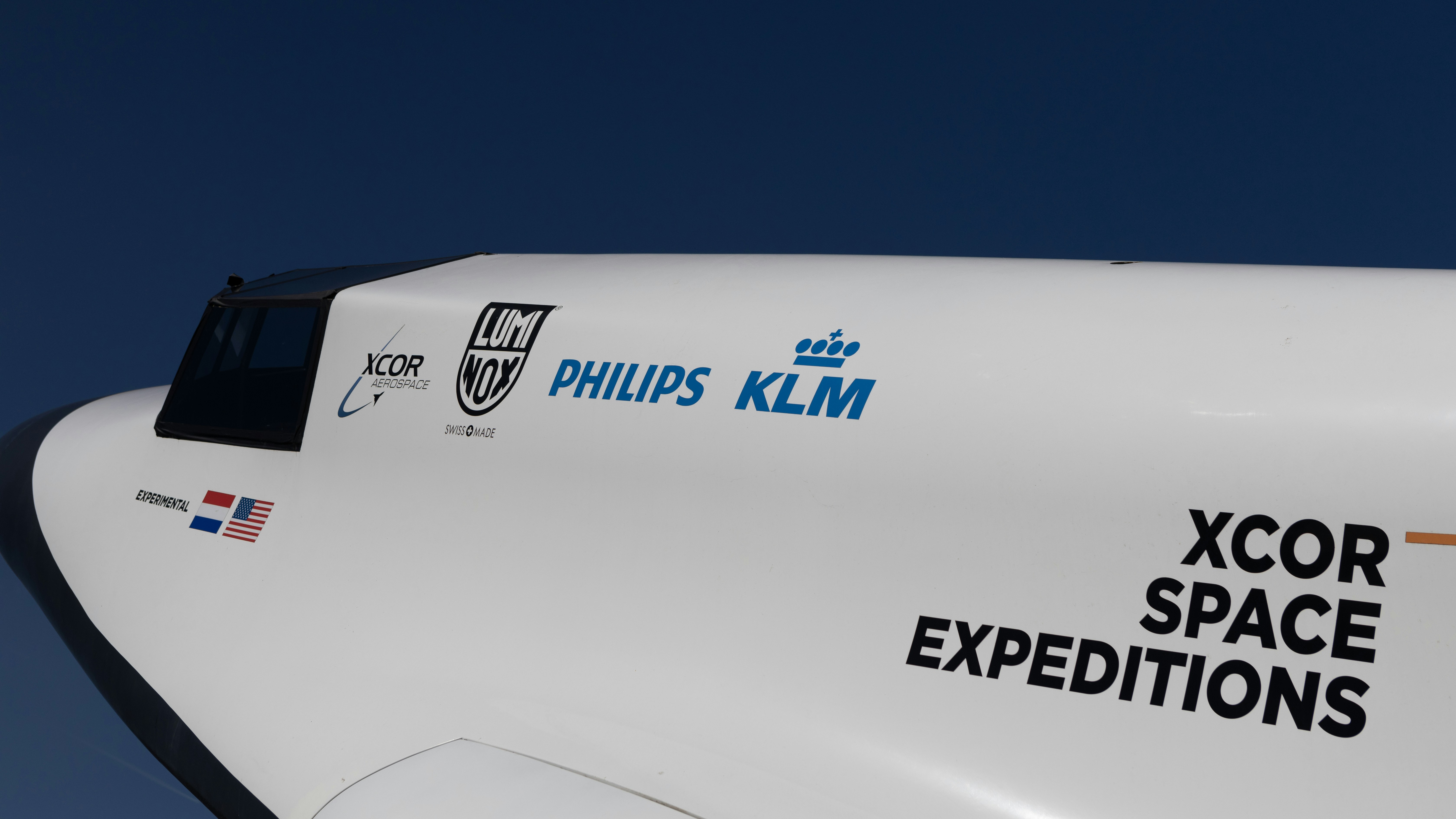 The sleek nose of a spacecraft featuring logos of XCOR, Philips, and KLM, set against a clear blue sky.