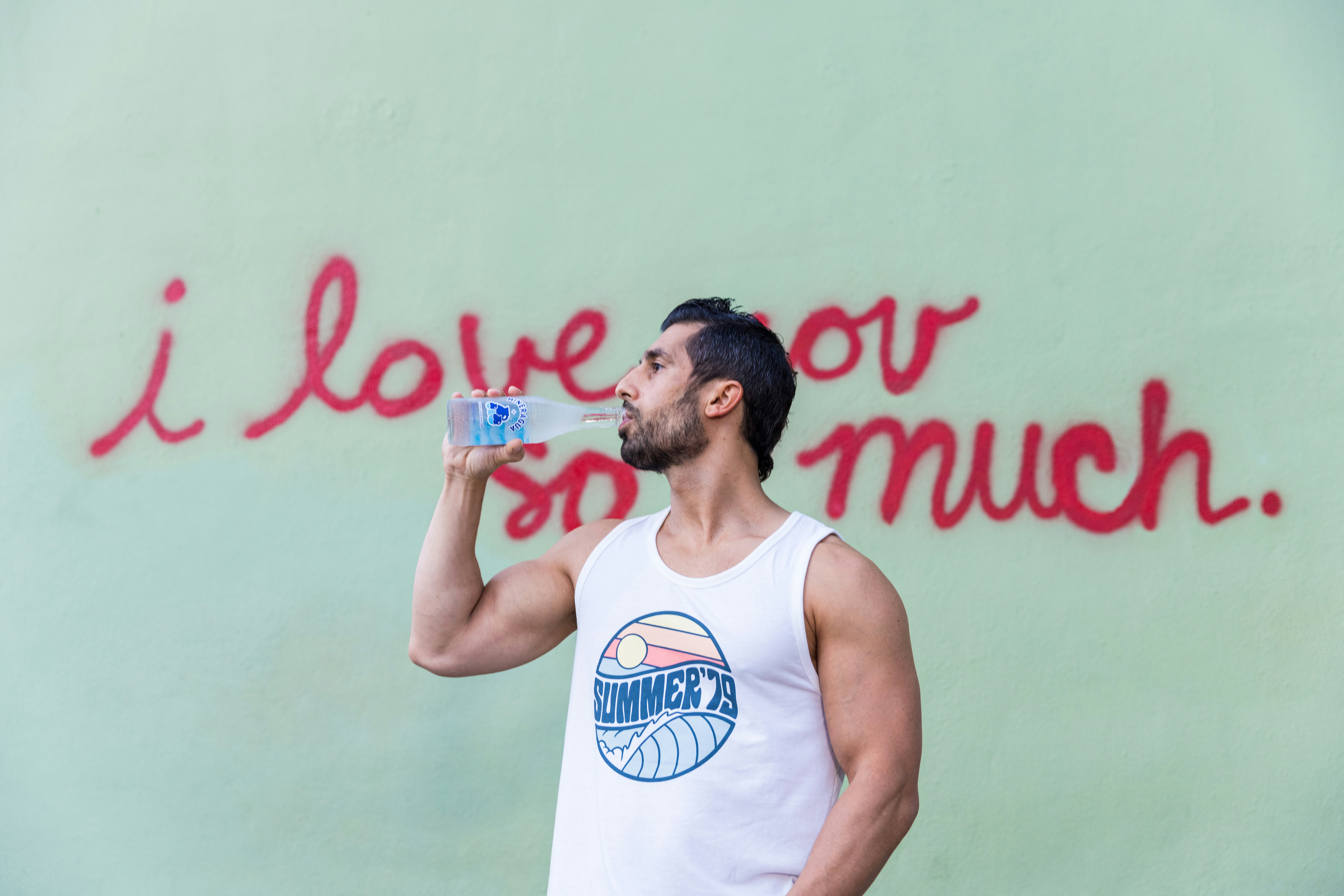 Man in a white tank top drinking water in front of green wall with red graffiti text.