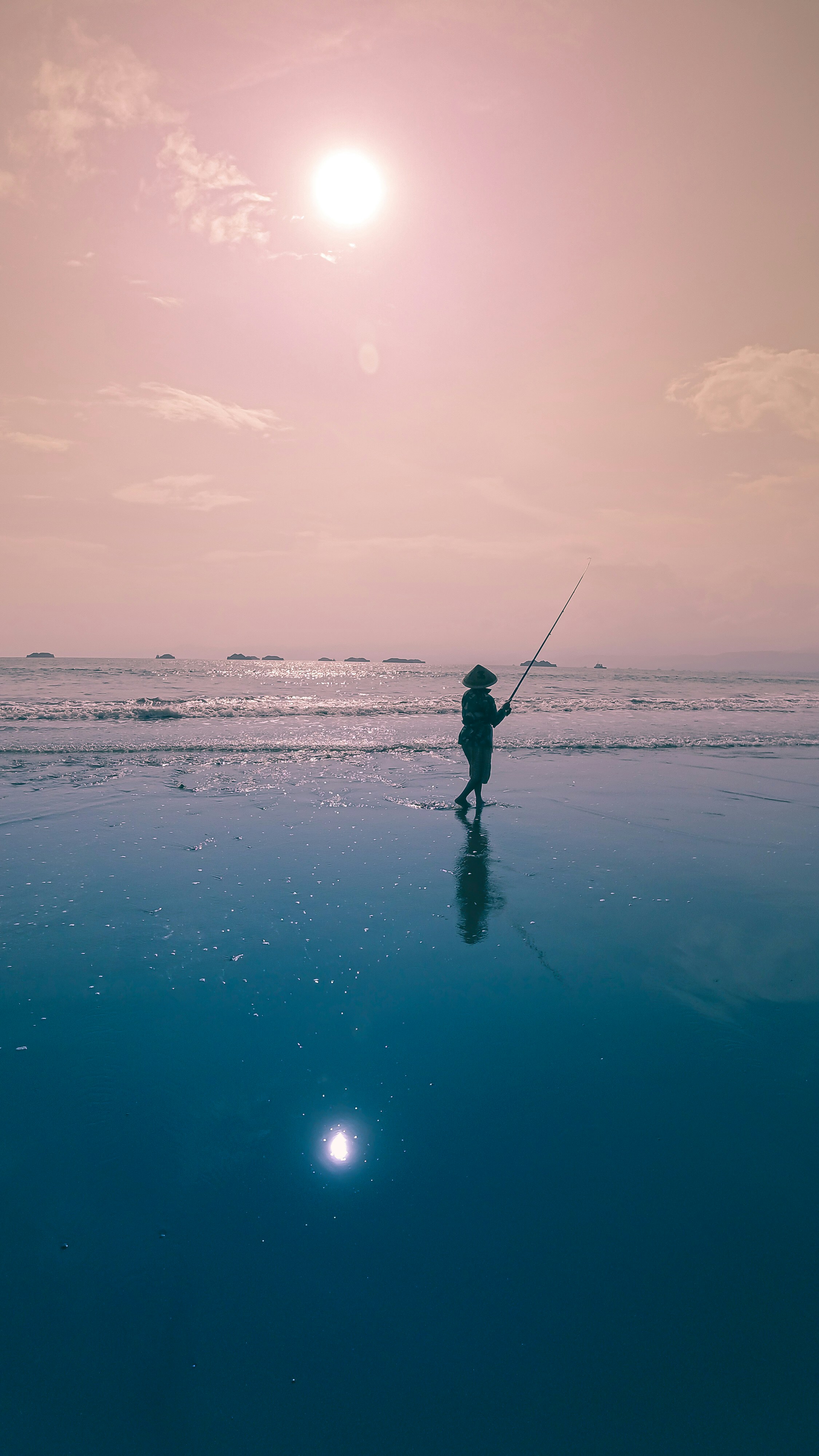 Silhouette of a fisherman casting a line on a reflective beach under a rising sun.