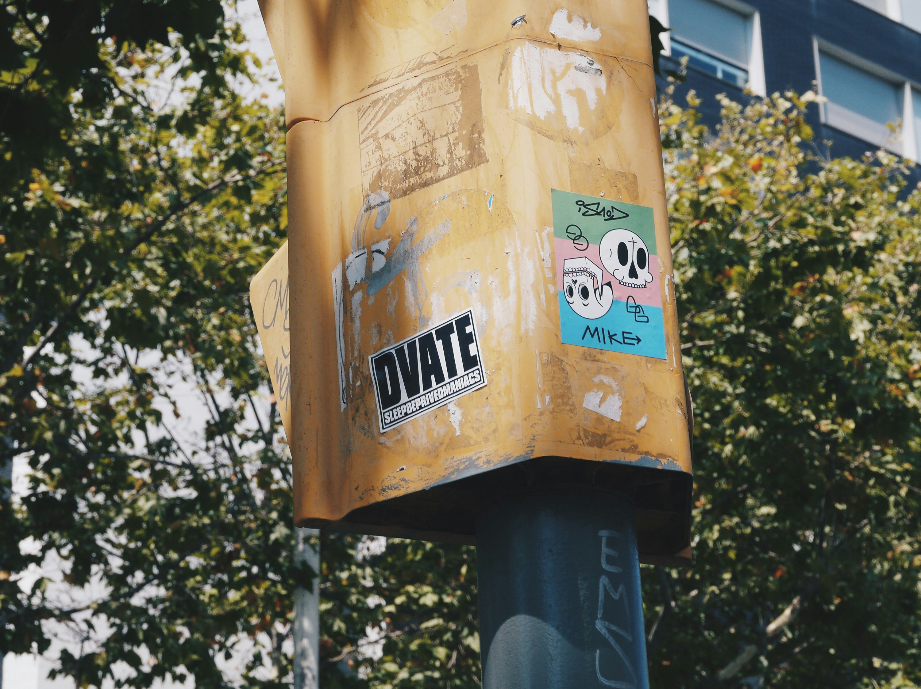 Close-up of a weathered yellow street lamp post covered in stickers and graffiti, with a leafy urban backdrop.