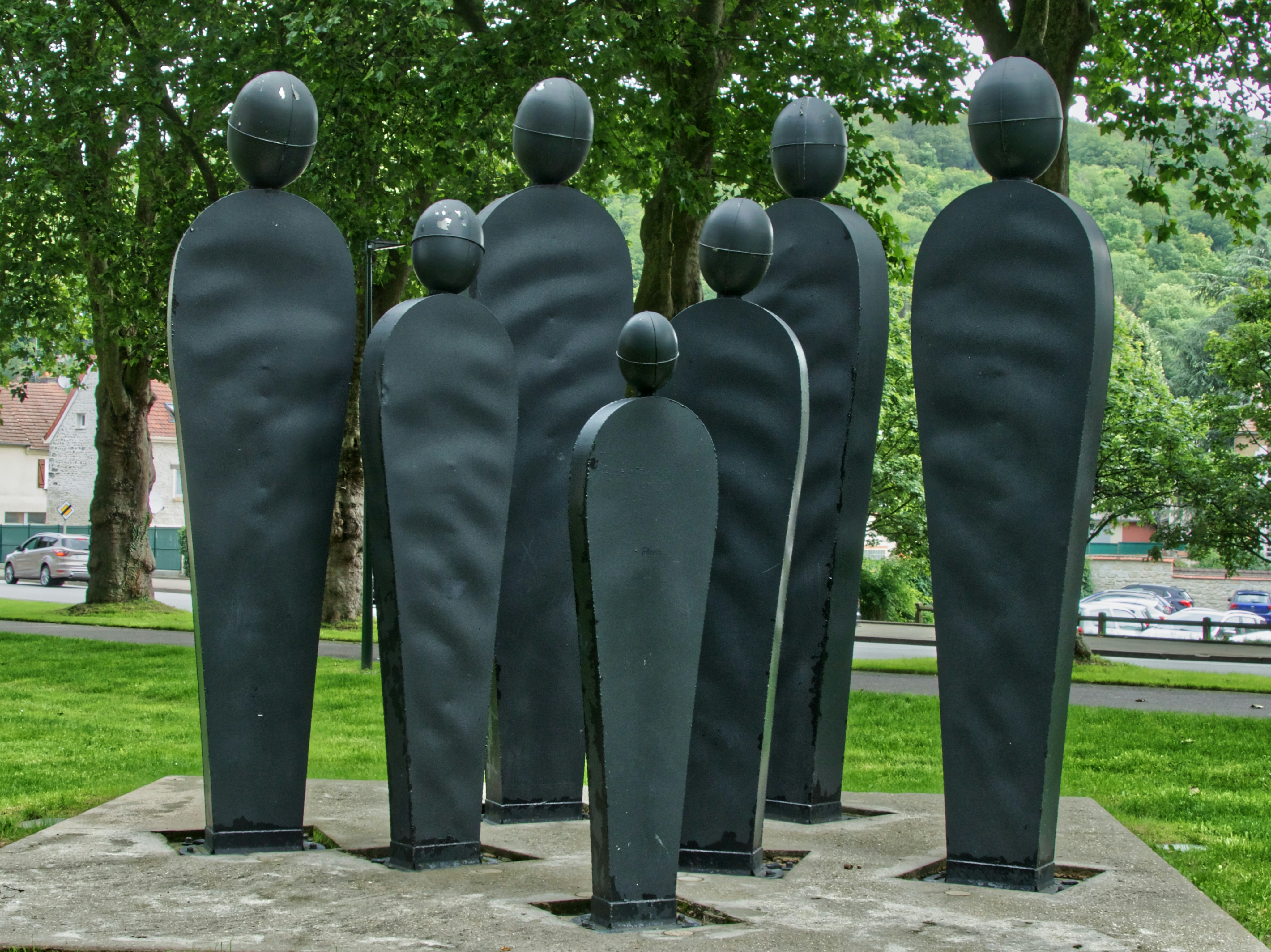 Abstract black metal sculptures resembling human figures stand in a park surrounded by lush green trees.
