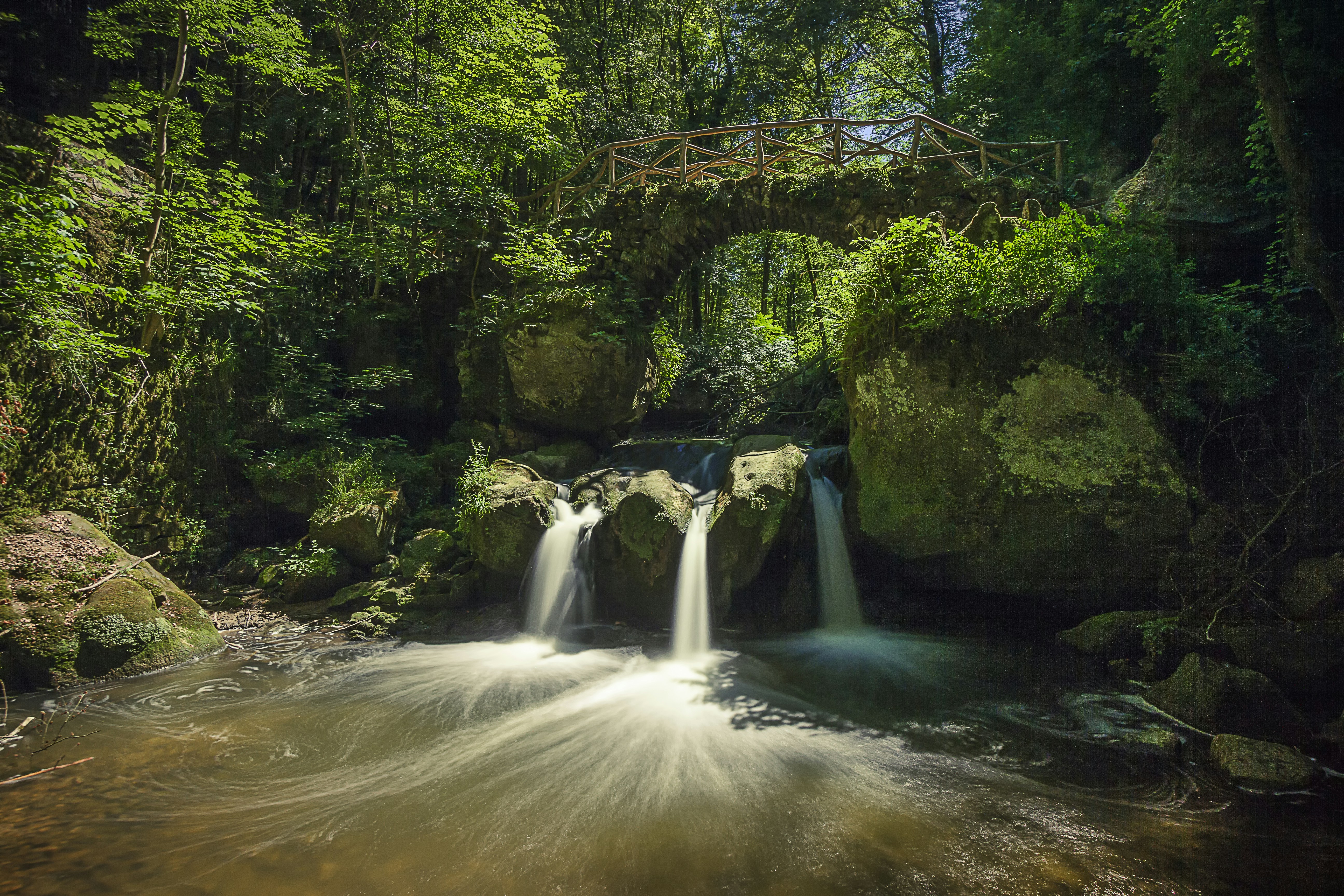 A serene waterfall cascading beneath a rustic stone bridge, surrounded by lush greenery and vibrant foliage.