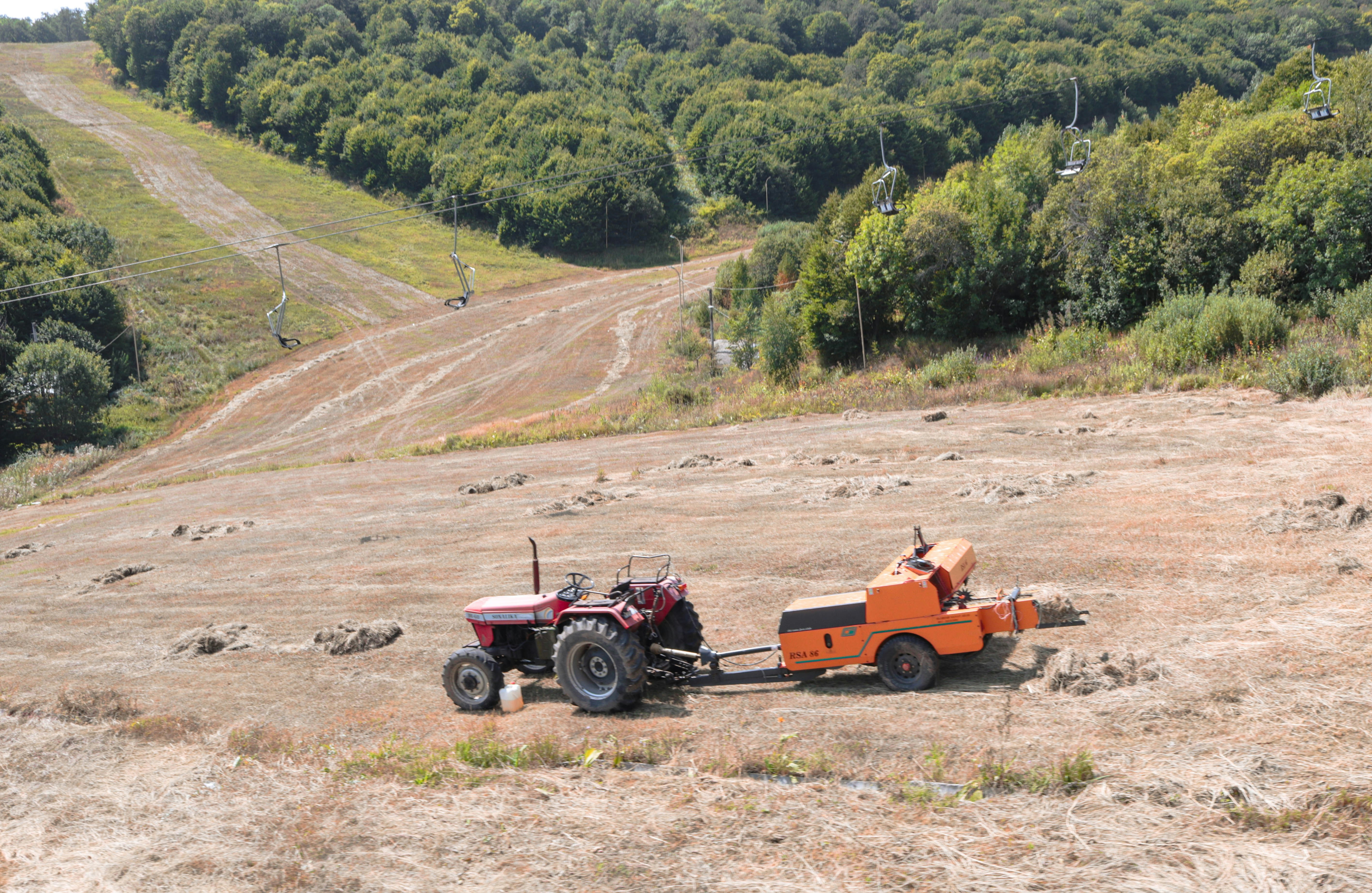 Red tractor towing an orange implement on a hillside, surrounded by lush green trees and ski lift chairs in the background.
