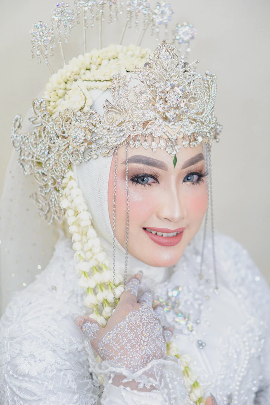 A smiling bride in an elaborate white headdress and jeweled crown, wearing delicate lace gloves and soft makeup. The background is plain and light, keeping focus on her face and ornate details.