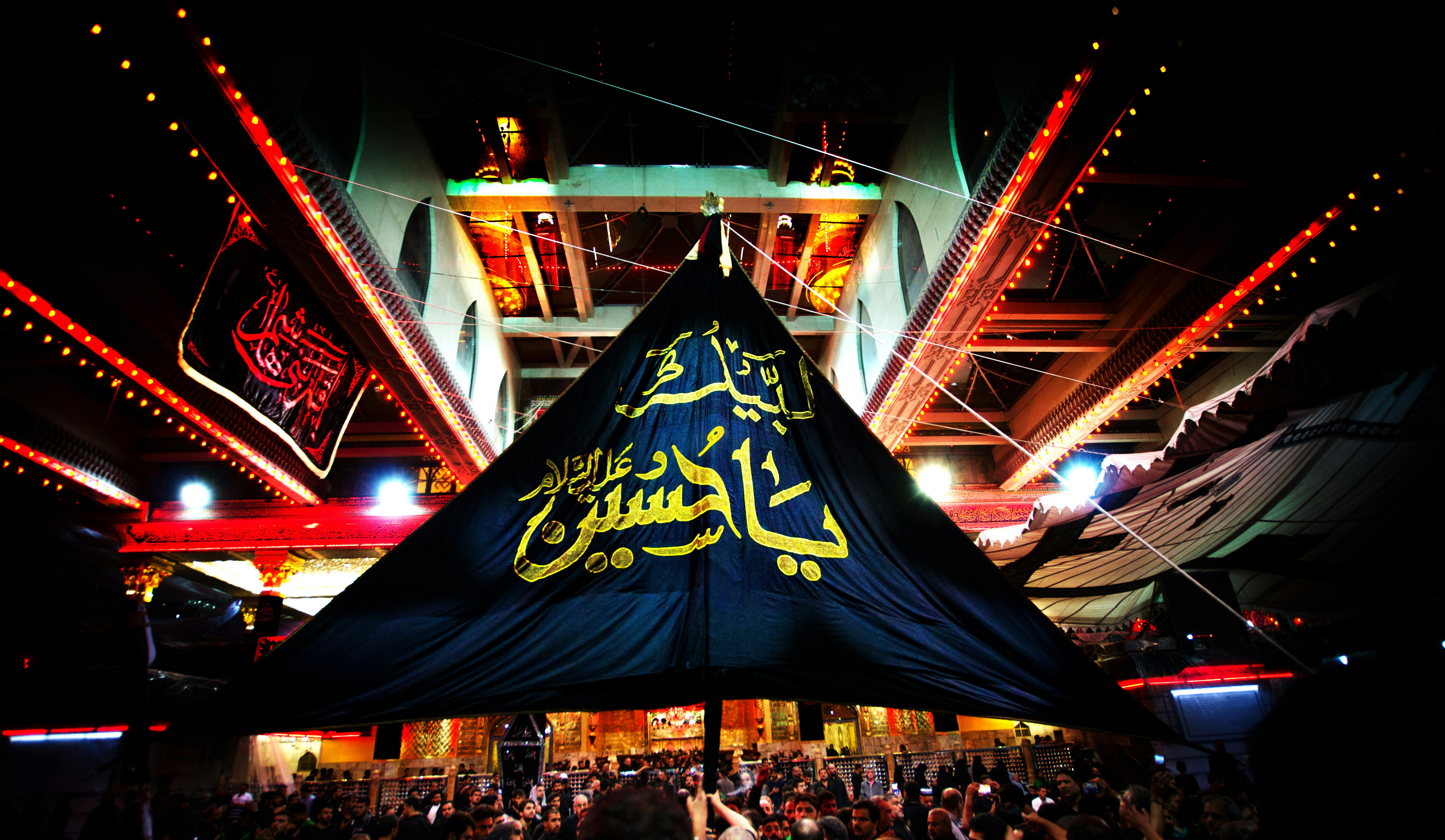 A vibrant mourning tent adorned with intricate calligraphy, set against a backdrop of illuminated decorations in a solemn gathering. The atmosphere reflects deep cultural significance.