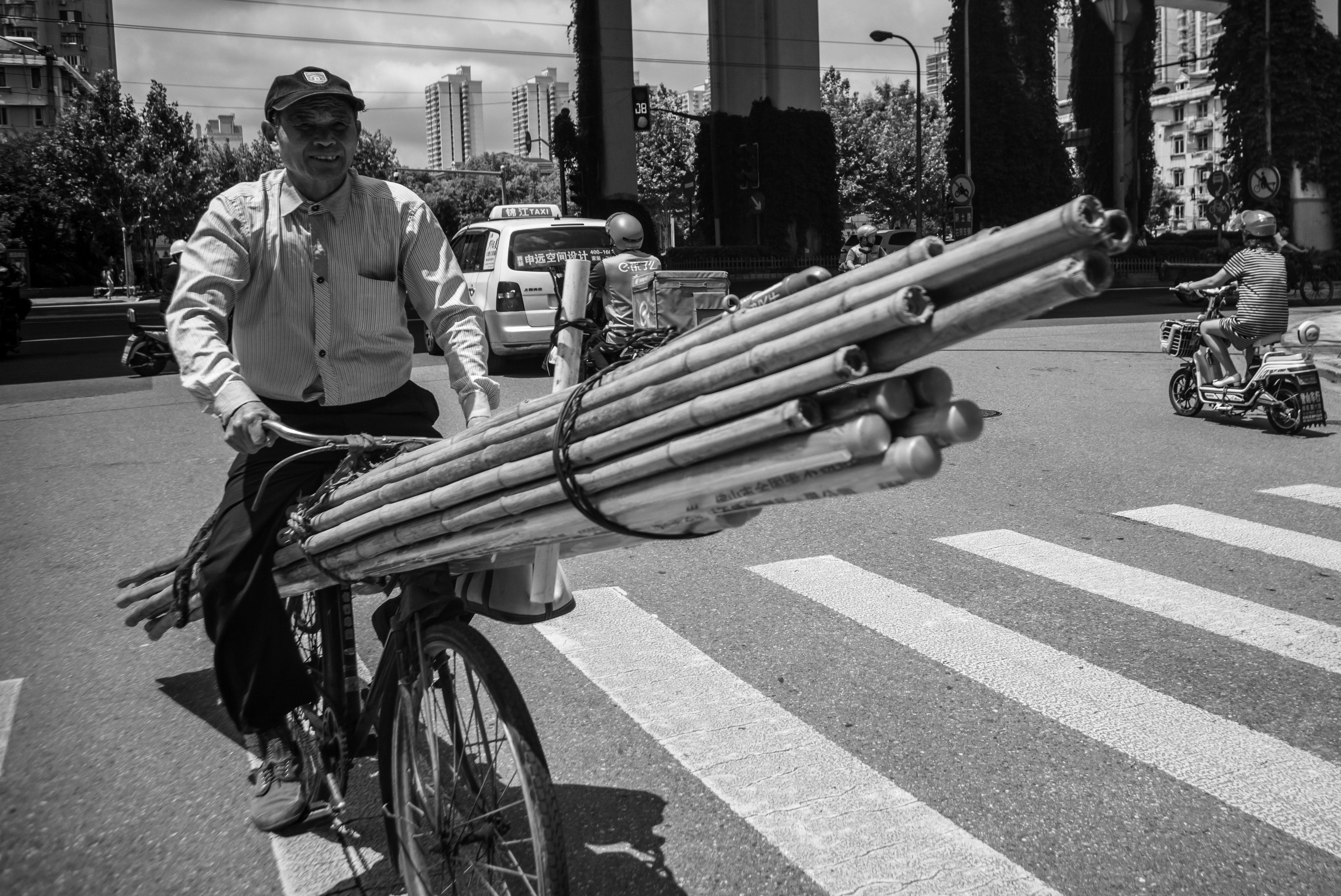 An elderly man balances a heavy load of bamboo poles on his bicycle while navigating a bustling city intersection.