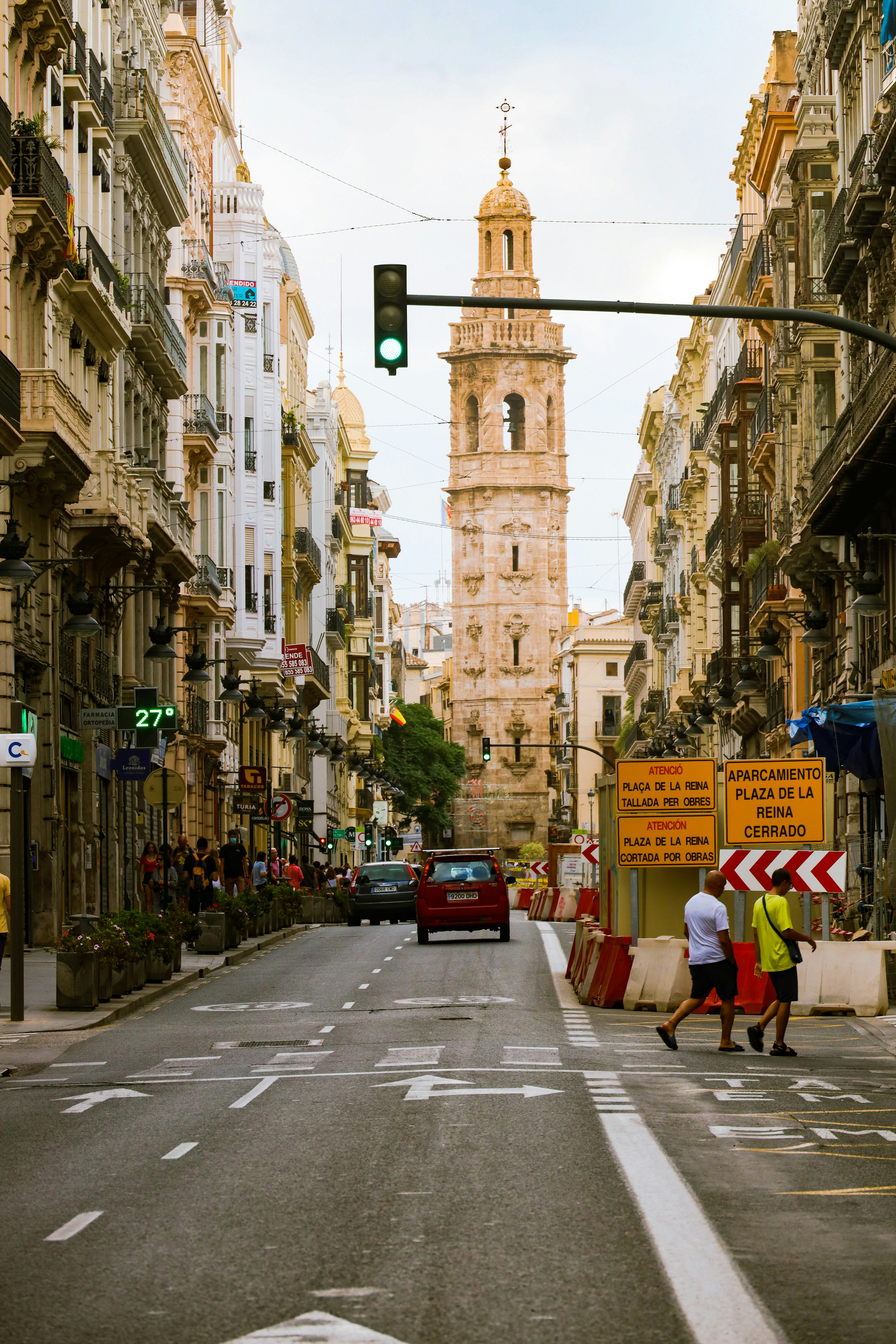 Historic clock tower stands tall at the end of a bustling street, framed by contemporary architecture and construction signs. 