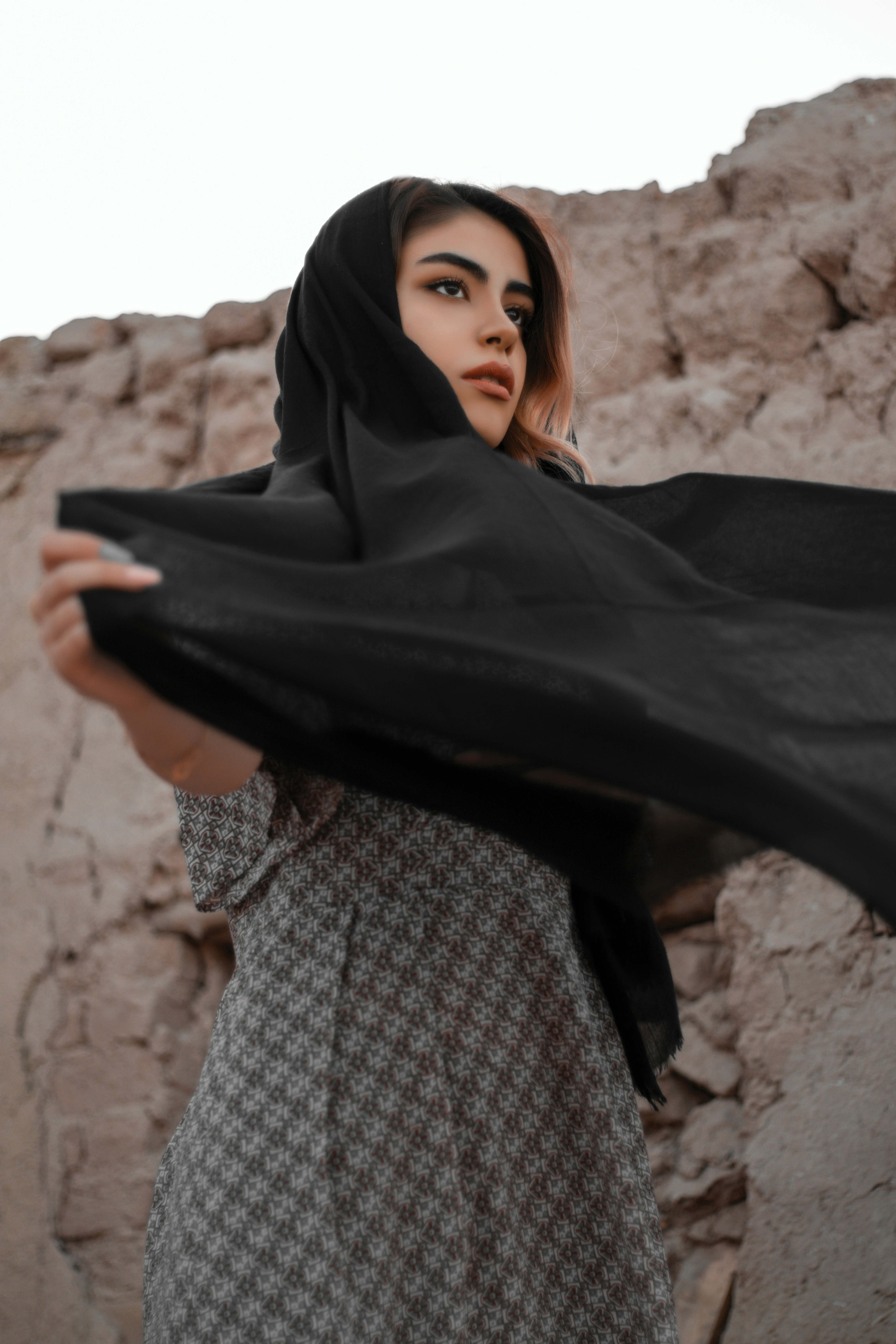 A young woman gracefully holds a flowing black scarf against a textured backdrop, embodying a blend of tradition and contemporary style.