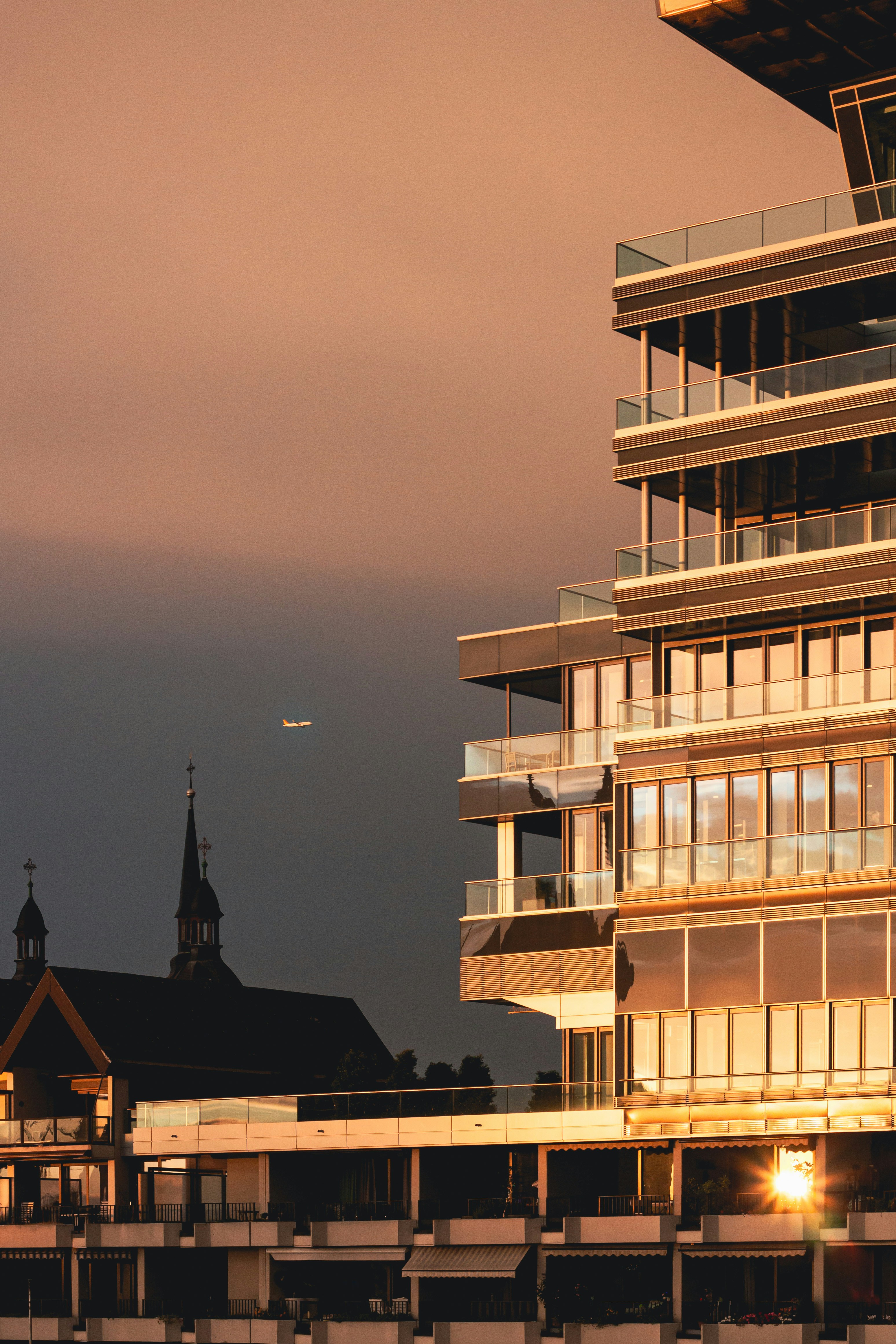 Modern architecture reflecting warm hues of sunset, with a distant airplane gliding through the sky. The scene captures a blend of urban life and natural beauty.