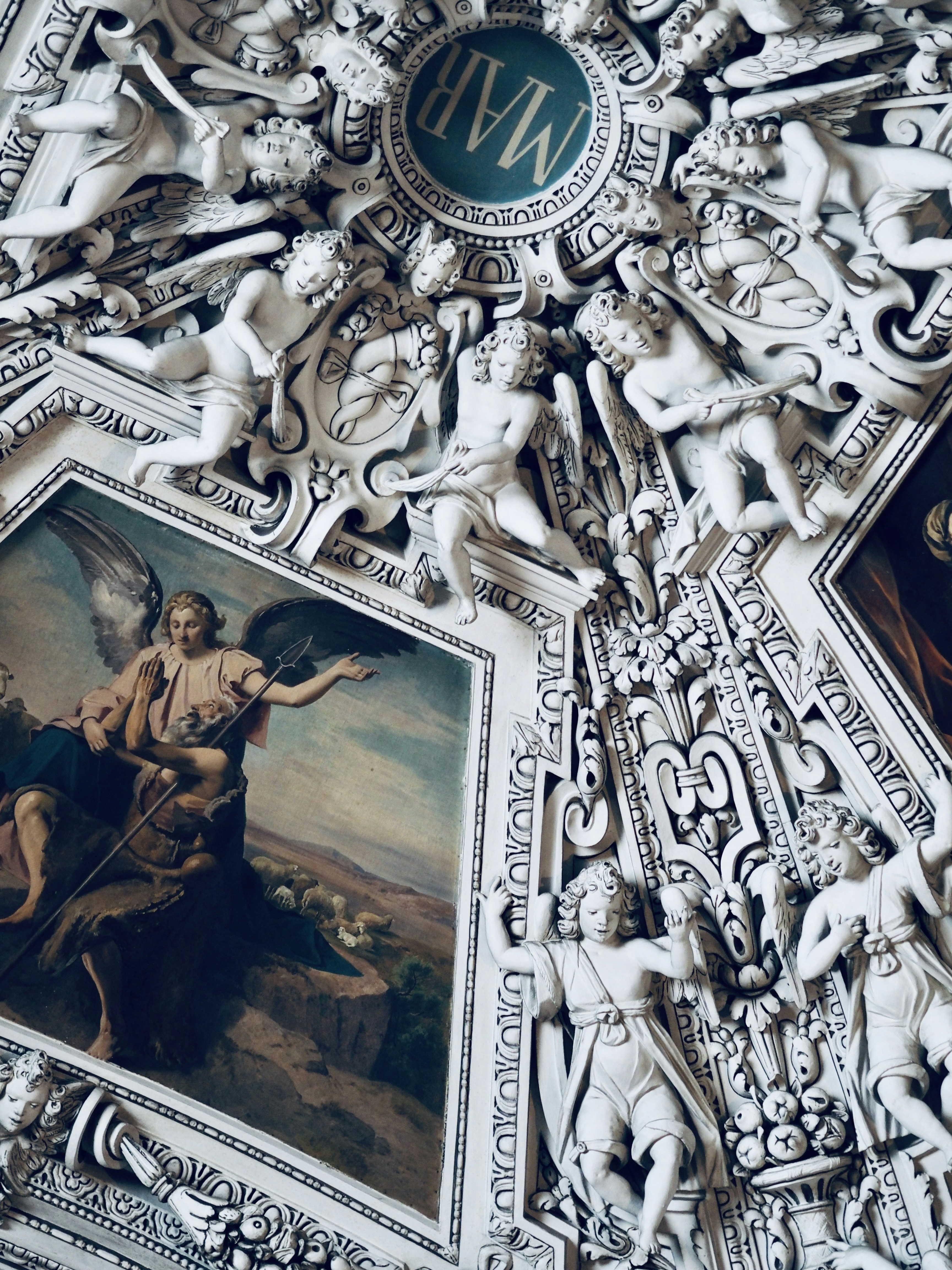 Intricate ceiling adorned with sculpted angels and a central painting, showcasing a blend of artistry and architectural elegance.