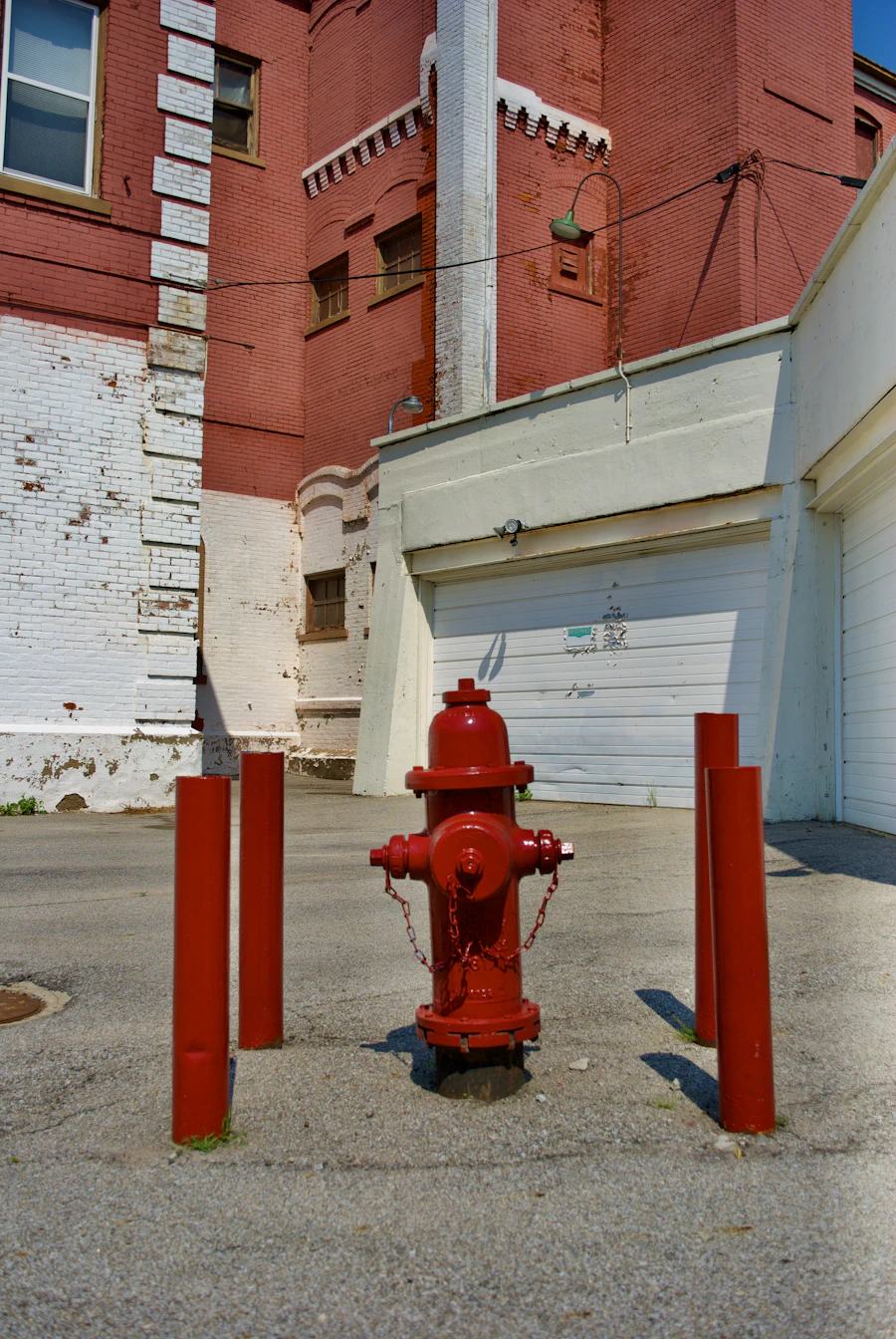 Bright red fire hydrant stands in front of a white garage door and weathered brick building, framed by four red posts. Sunlit urban alley with peeling paint and utility lines overhead.