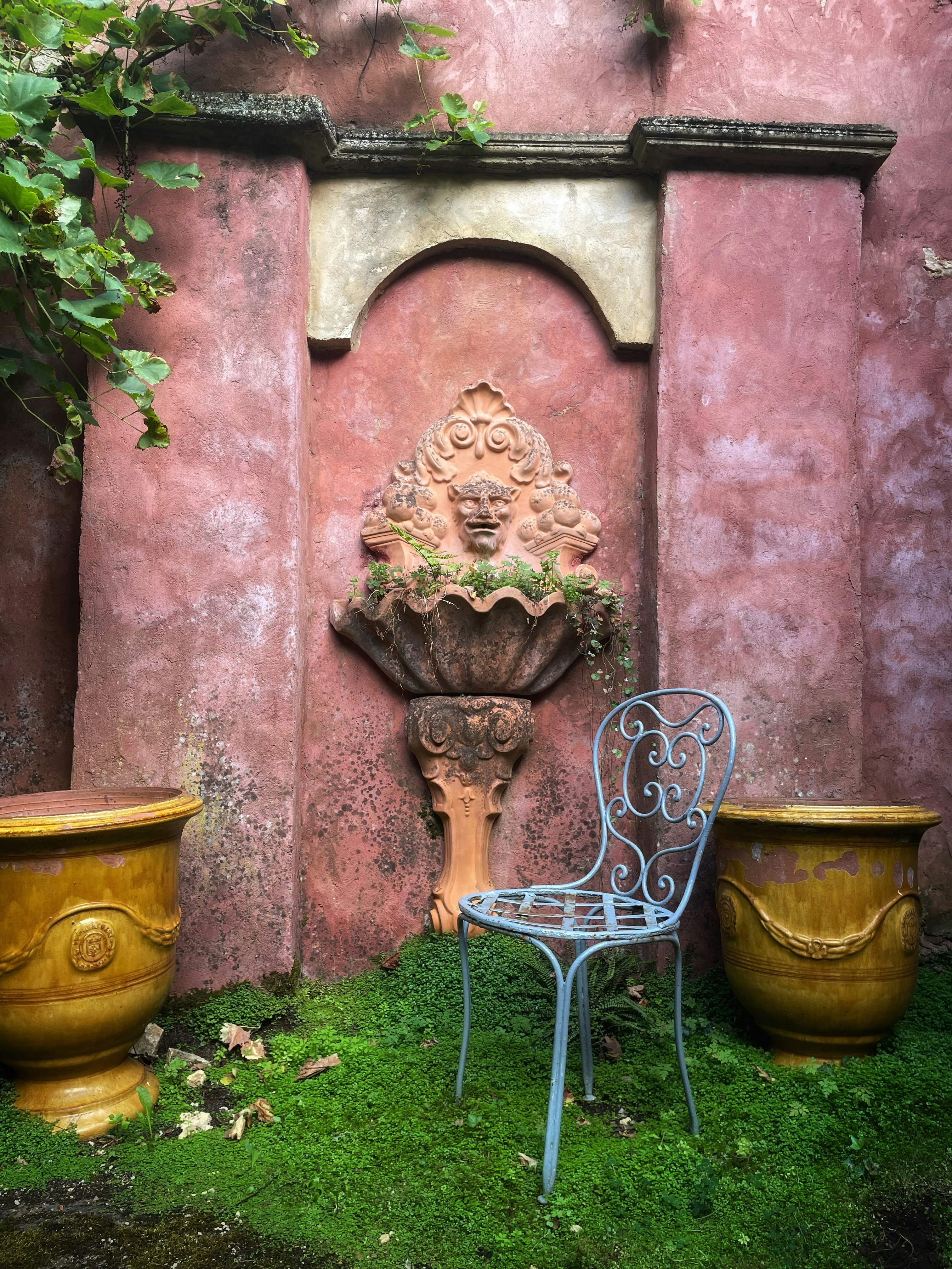 A delicate blue chair rests beside ornate planters, framed by a weathered pink wall adorned with a sculpted fountain. Lush greenery peeks through, hinting at nature's reclaiming touch.