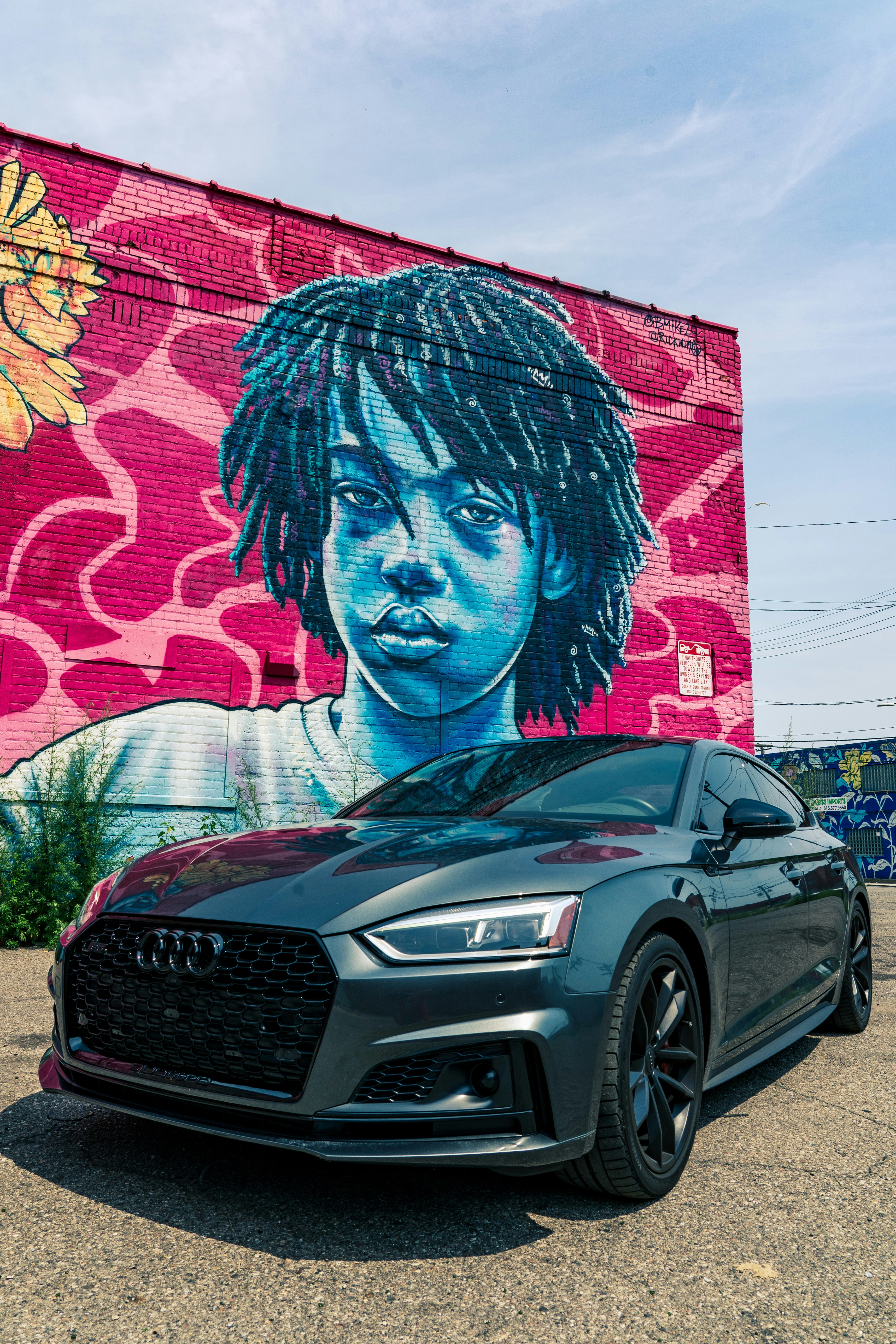 A sleek Audi A5 positioned in front of a vibrant mural depicting a young person with striking blue skin and intricate hair. The urban backdrop enhances the car's modern design.