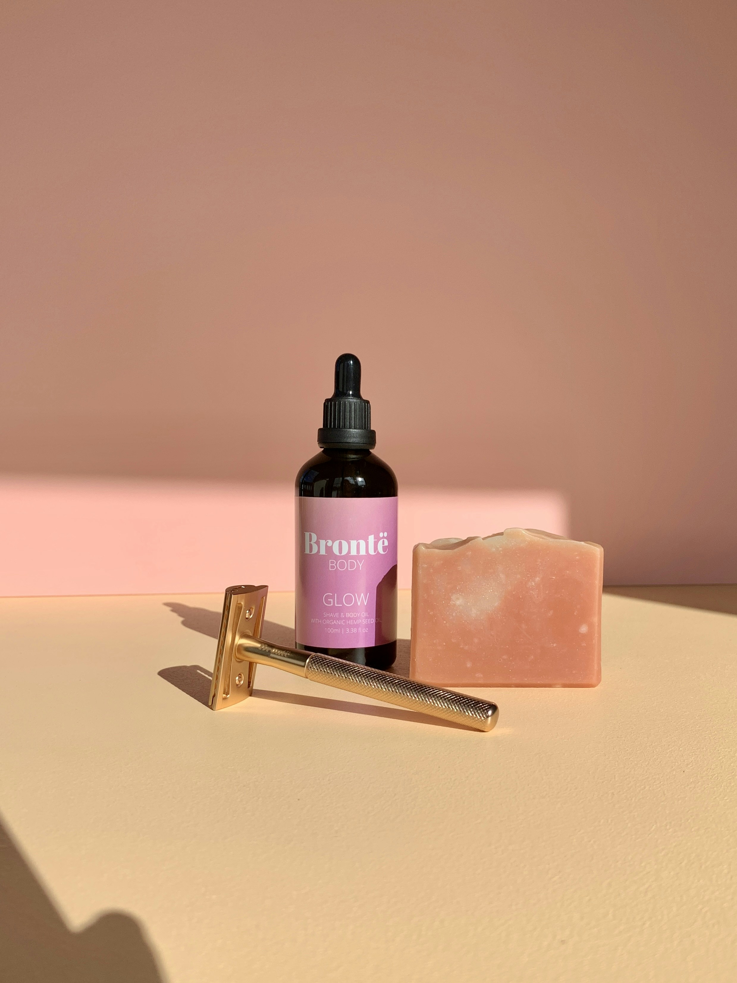 A gold safety razor, a bottle of Brontë body oil, and a pink soap bar arranged artistically against a soft peach background.