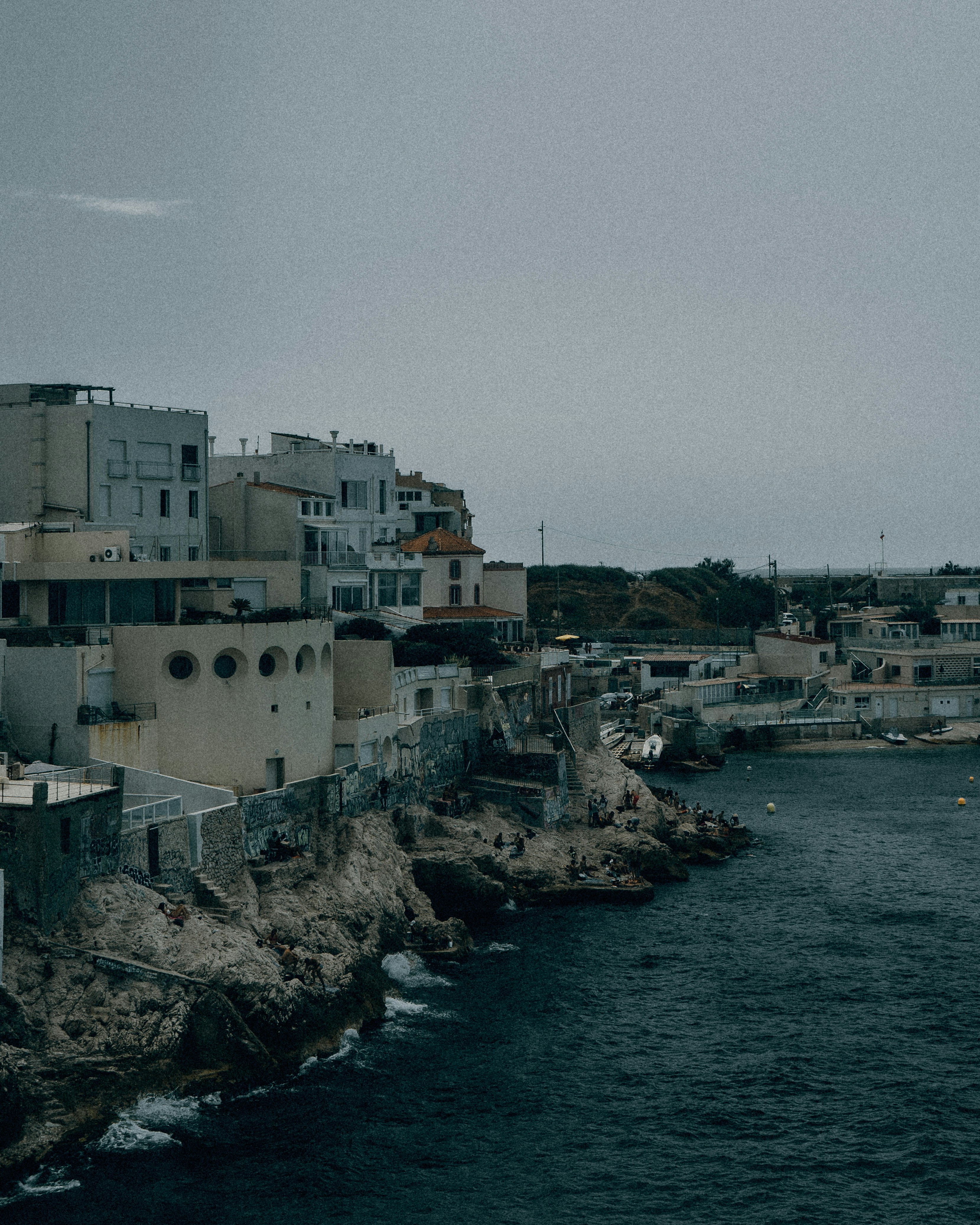 Coastal village with white buildings lining a rocky shore, overlooking a tranquil harbor under a cloudy sky.