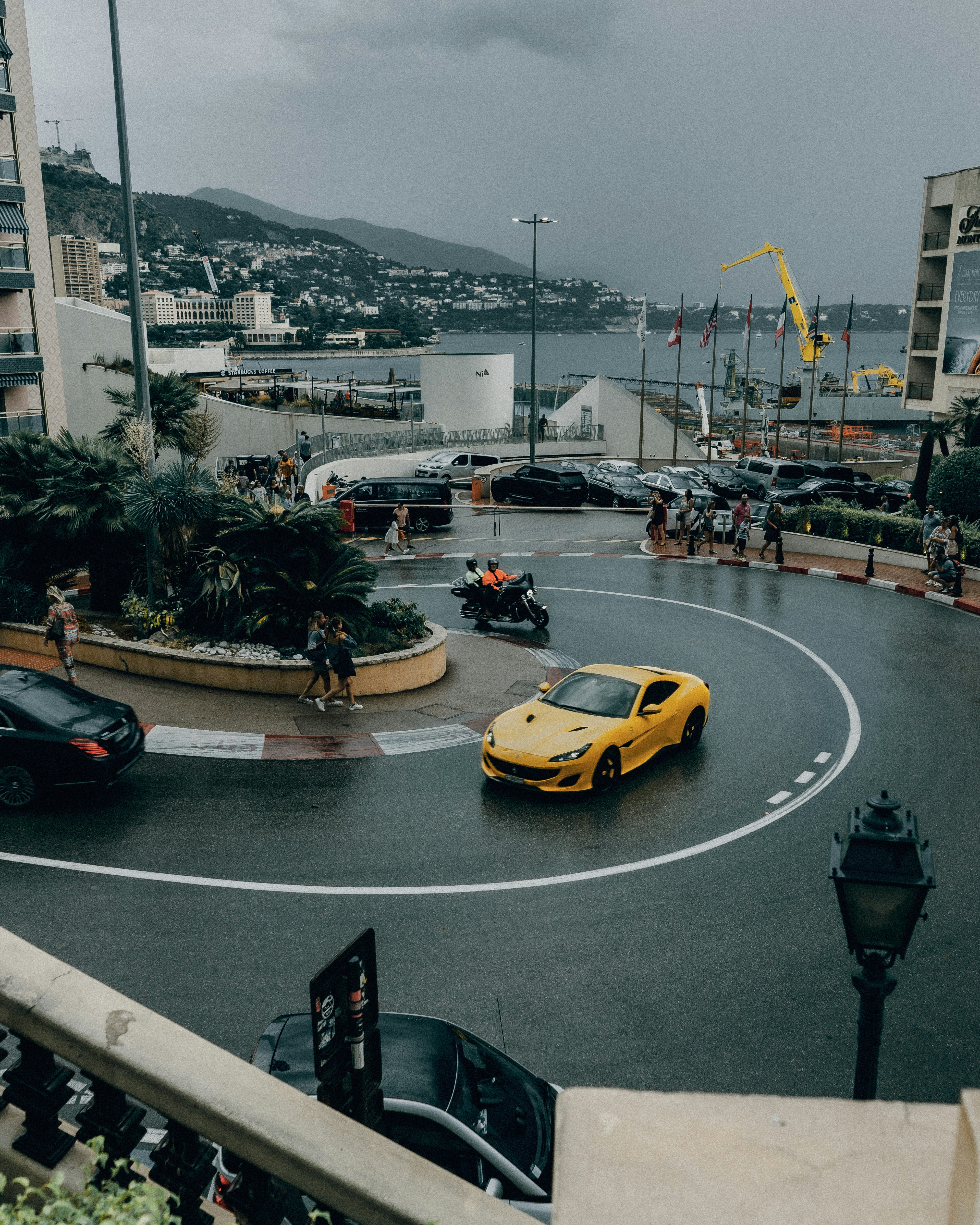 A yellow sports car navigates a winding road in Monaco, surrounded by lush greenery and bustling pedestrians. The dramatic sky hints at an impending storm.