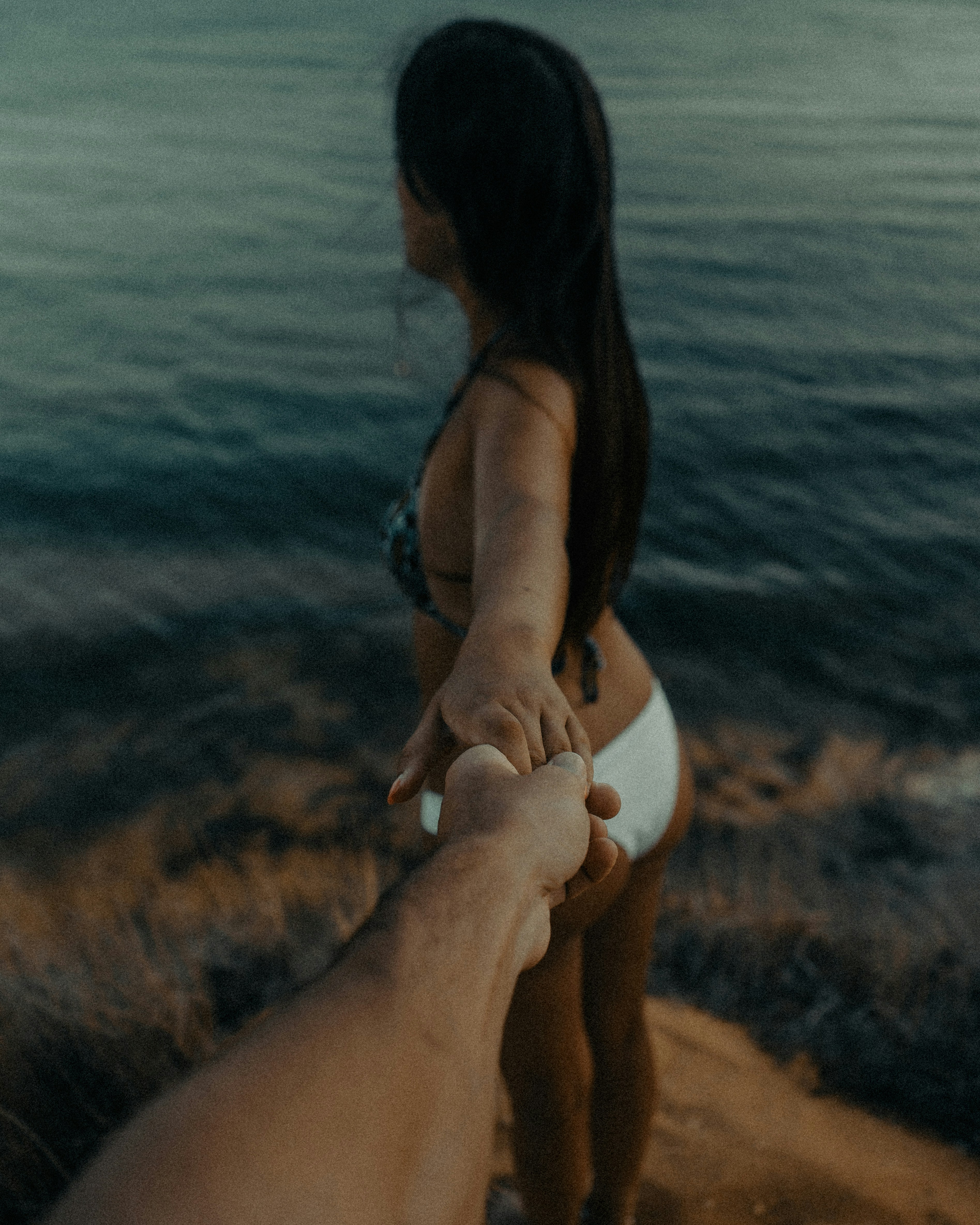 A woman in a bikini reaches back to hold hands with a partner, set against a tranquil water backdrop during golden hour.