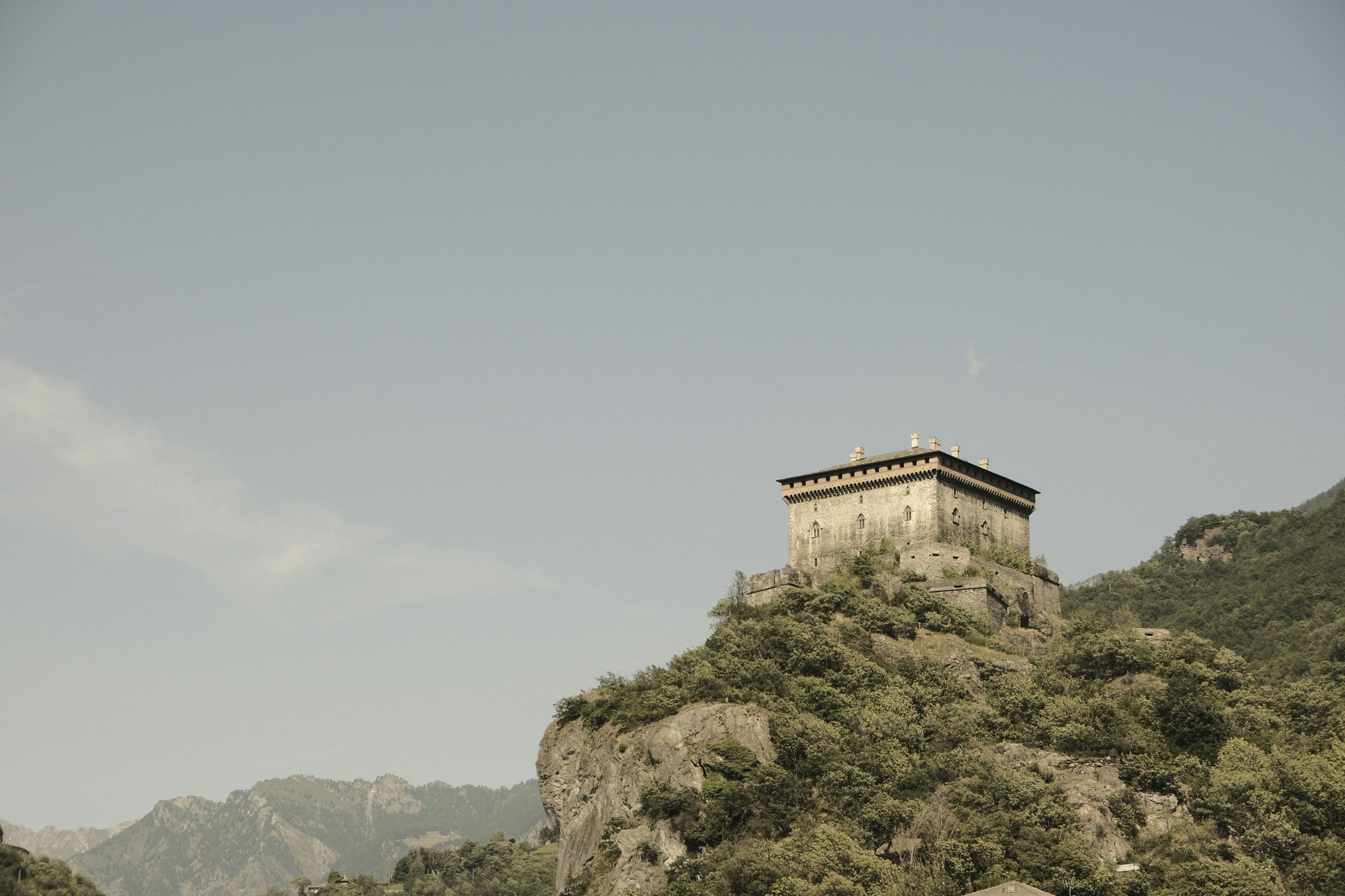 Historic fortress perched atop a rocky outcrop, surrounded by lush greenery and distant mountains under a clear sky.