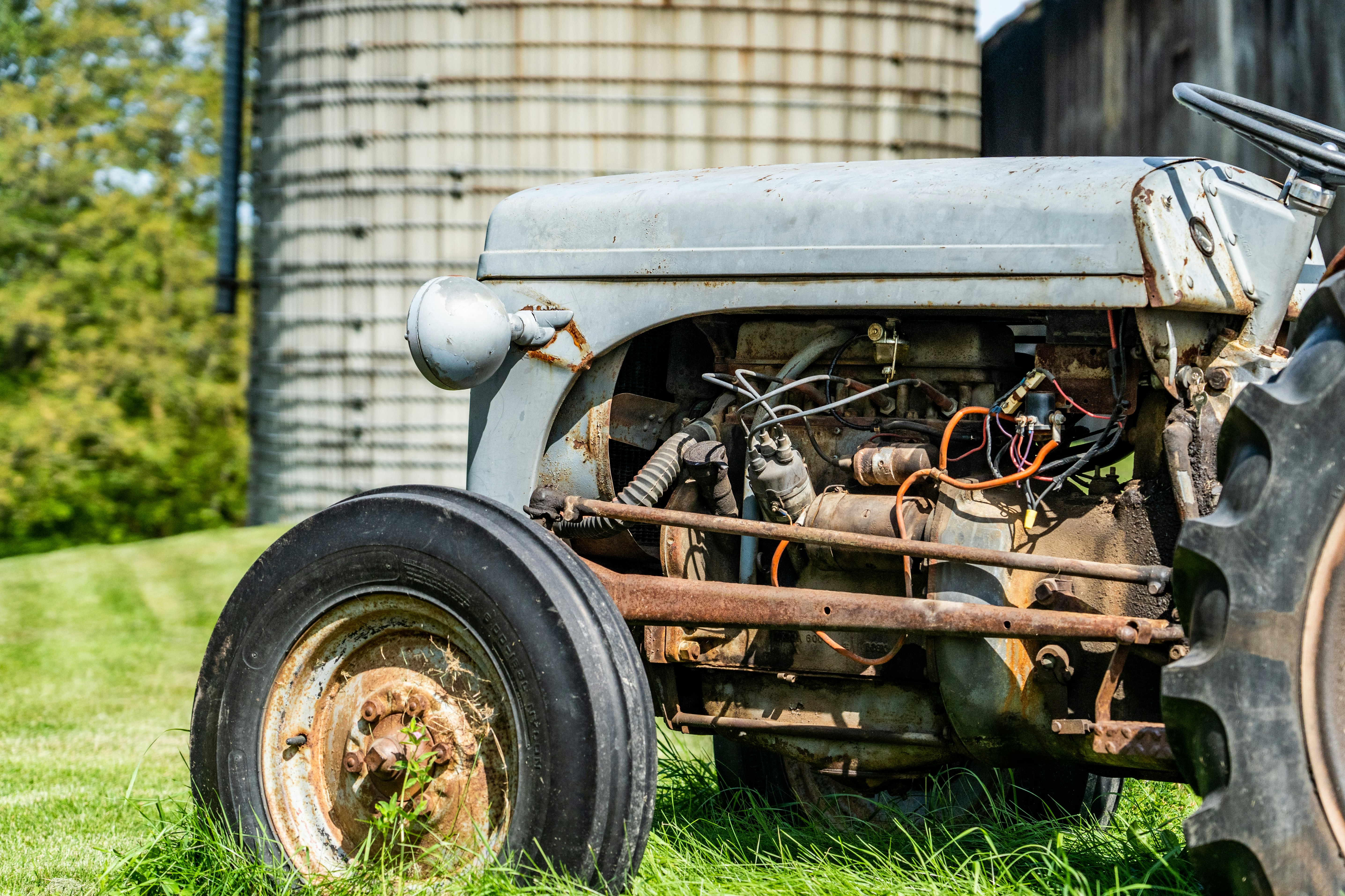 Close-up of an aged tractor revealing intricate engine details against a backdrop of a silos and greenery.