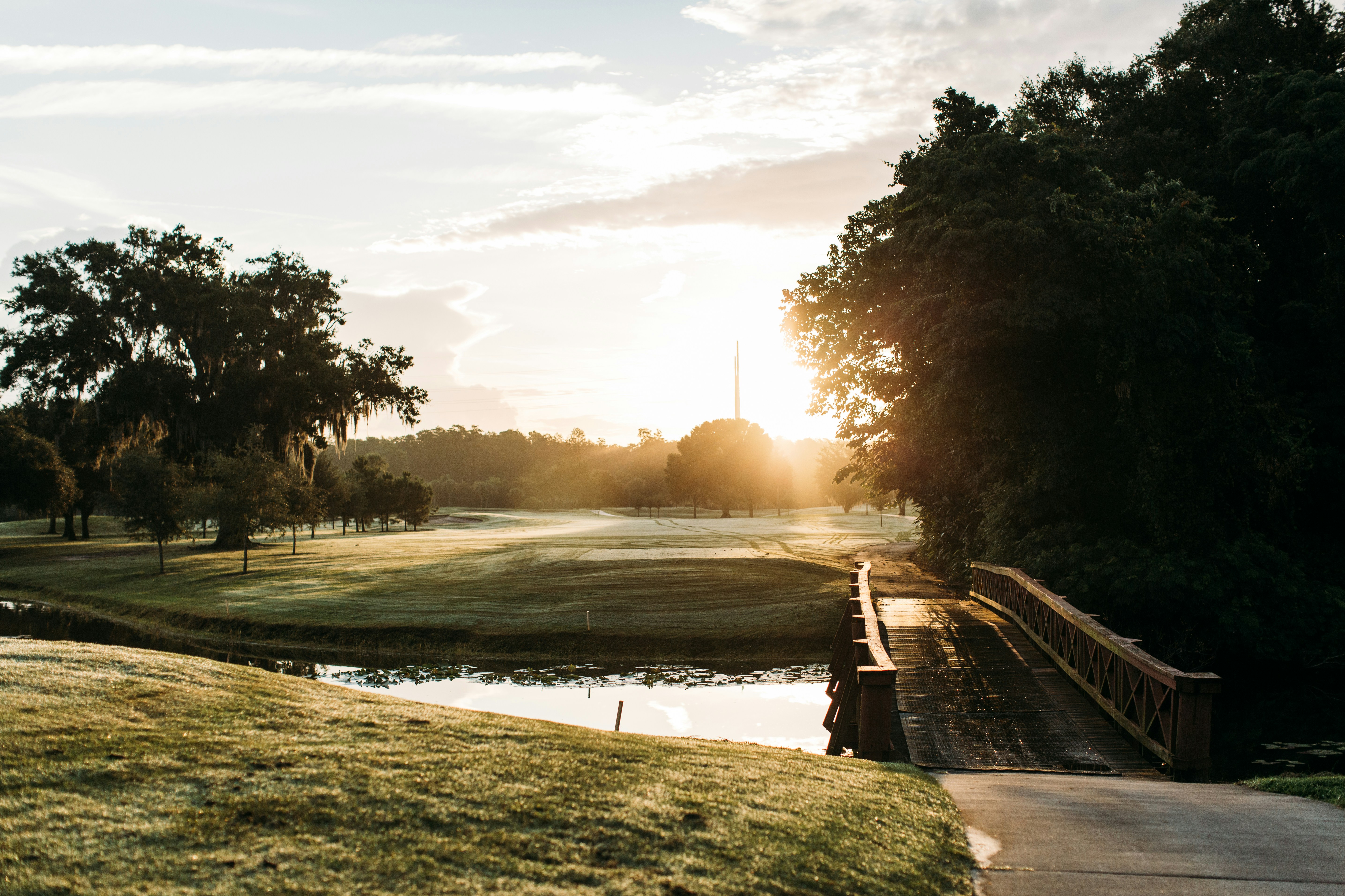 Sun rising over a golf course, illuminating a wooden bridge and surrounding greenery.