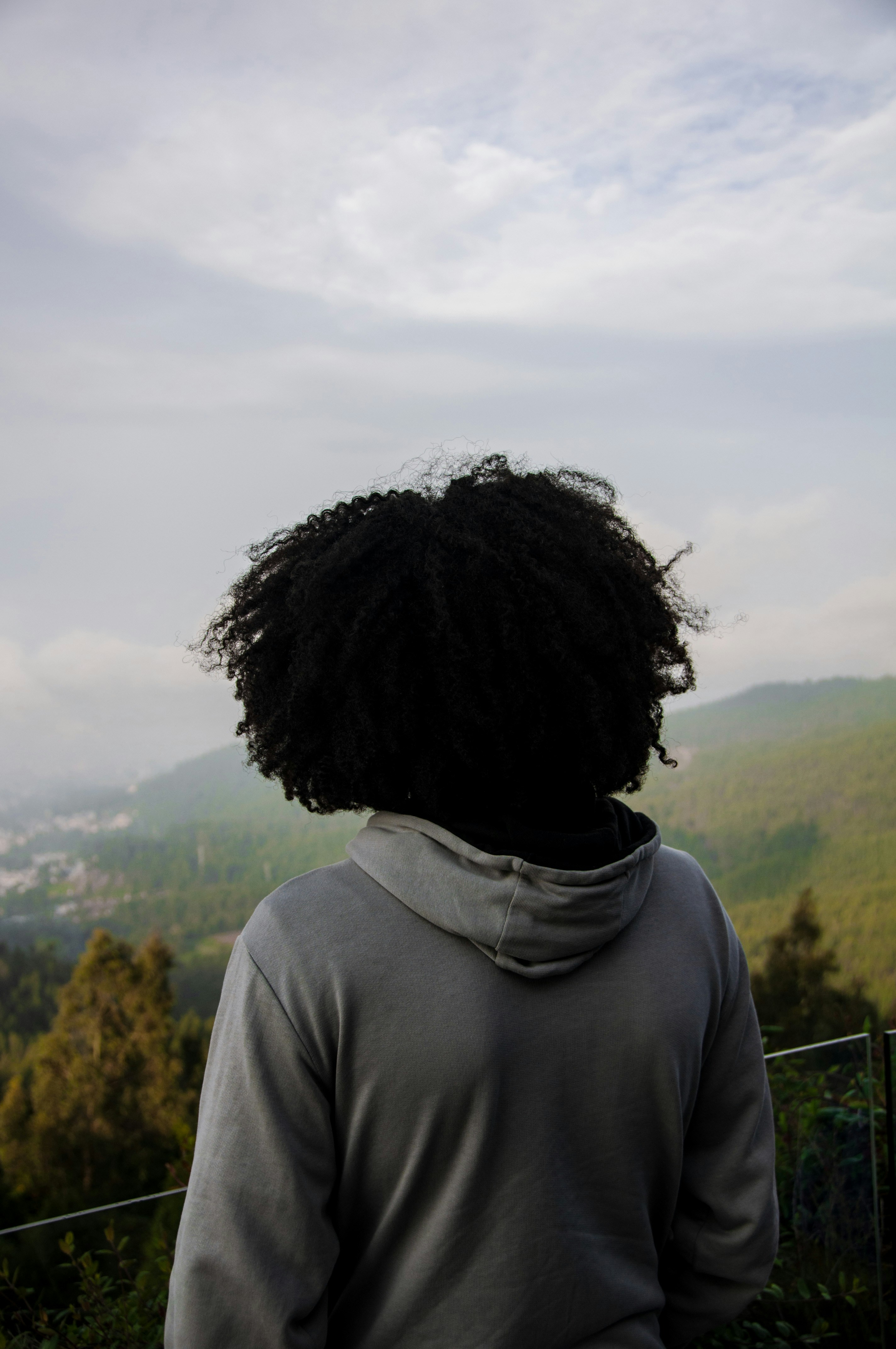 Individual with curly hair gazing over a lush landscape, enveloped in a serene atmosphere. The scene captures a moment of reflection against a backdrop of rolling hills.