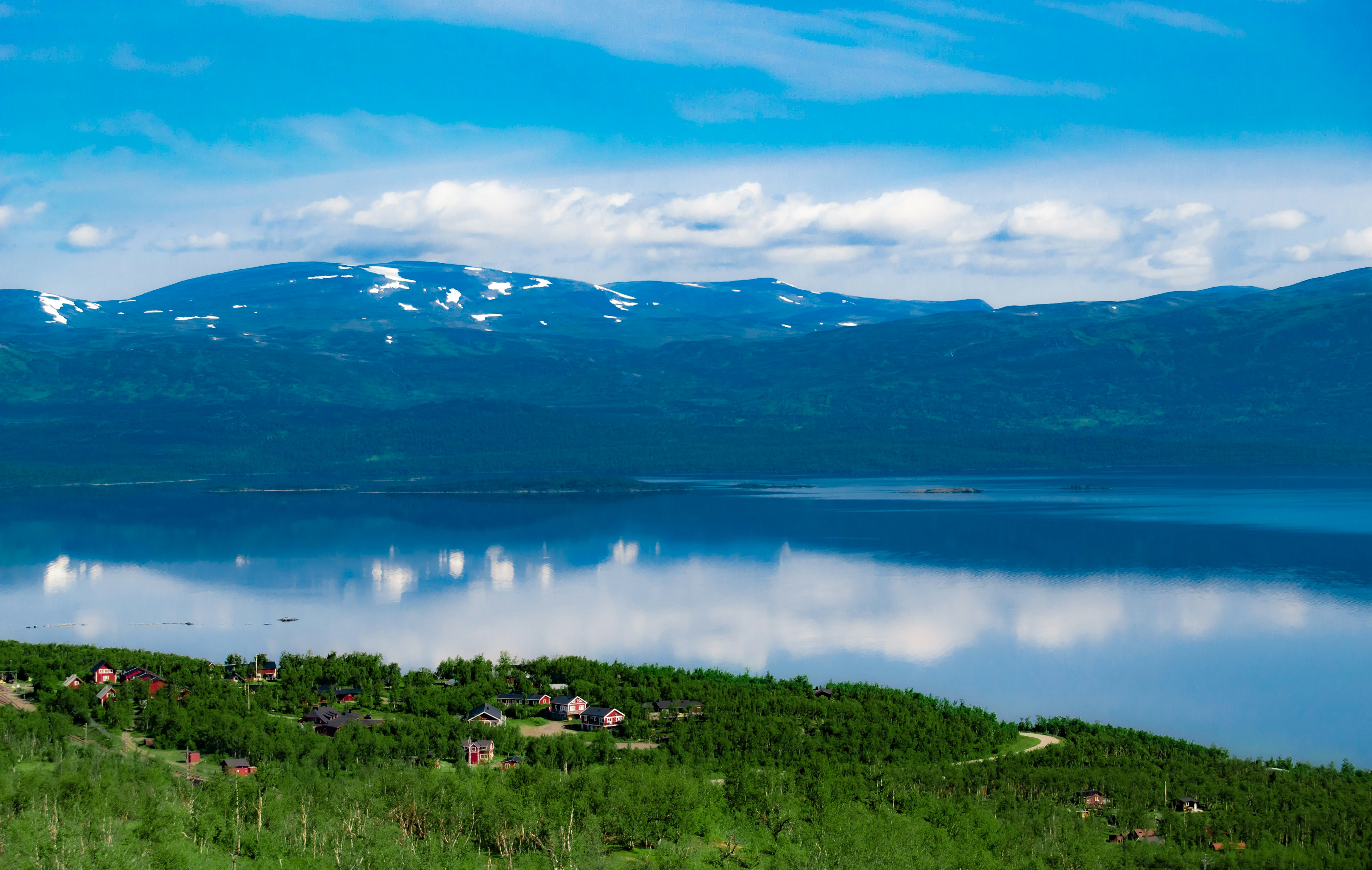 Lush green hills dotted with quaint houses reflect in the calm waters of a fjord, framed by distant snow-capped mountains under a clear blue sky.