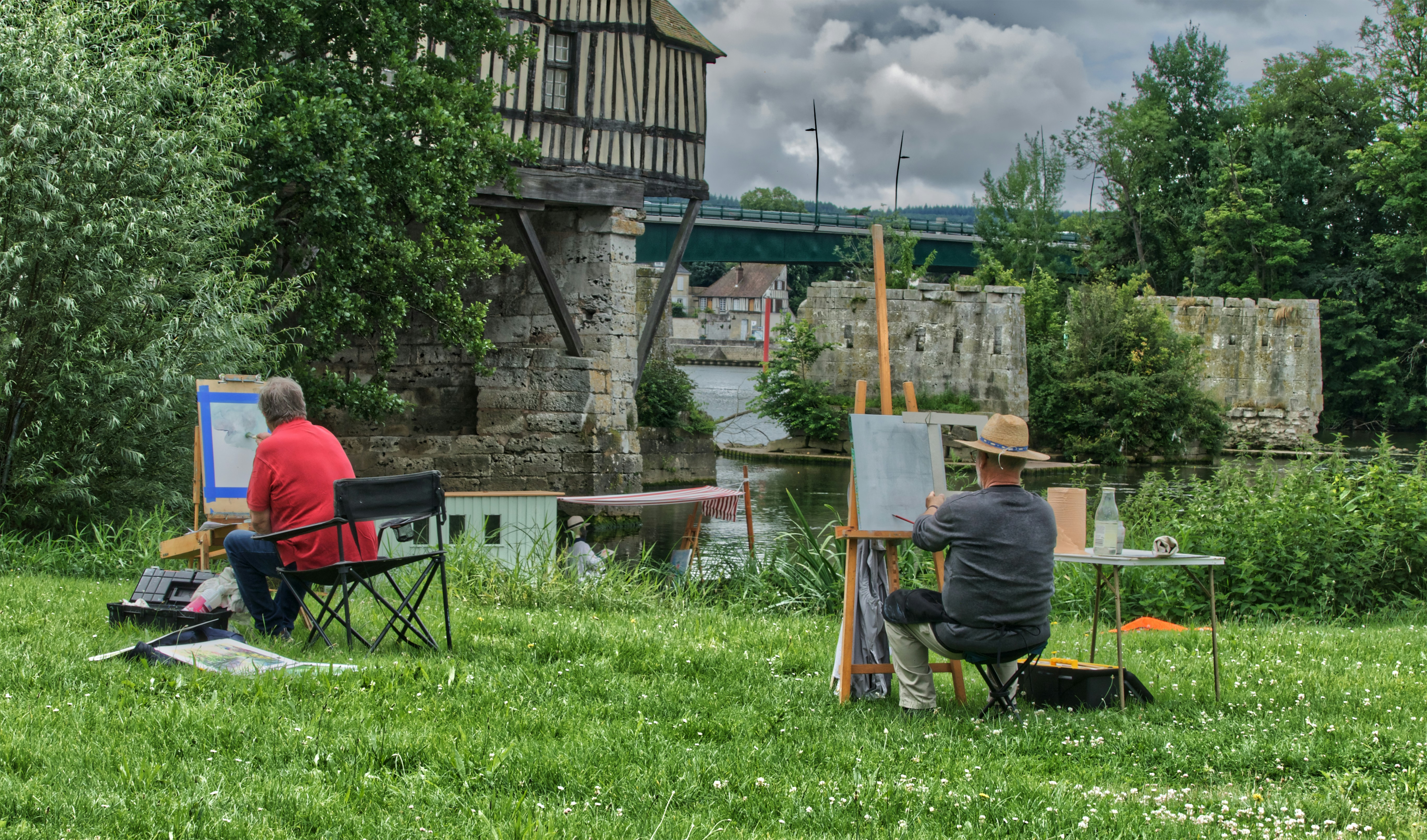 Artists painting by the river near historic stone structures and lush greenery.