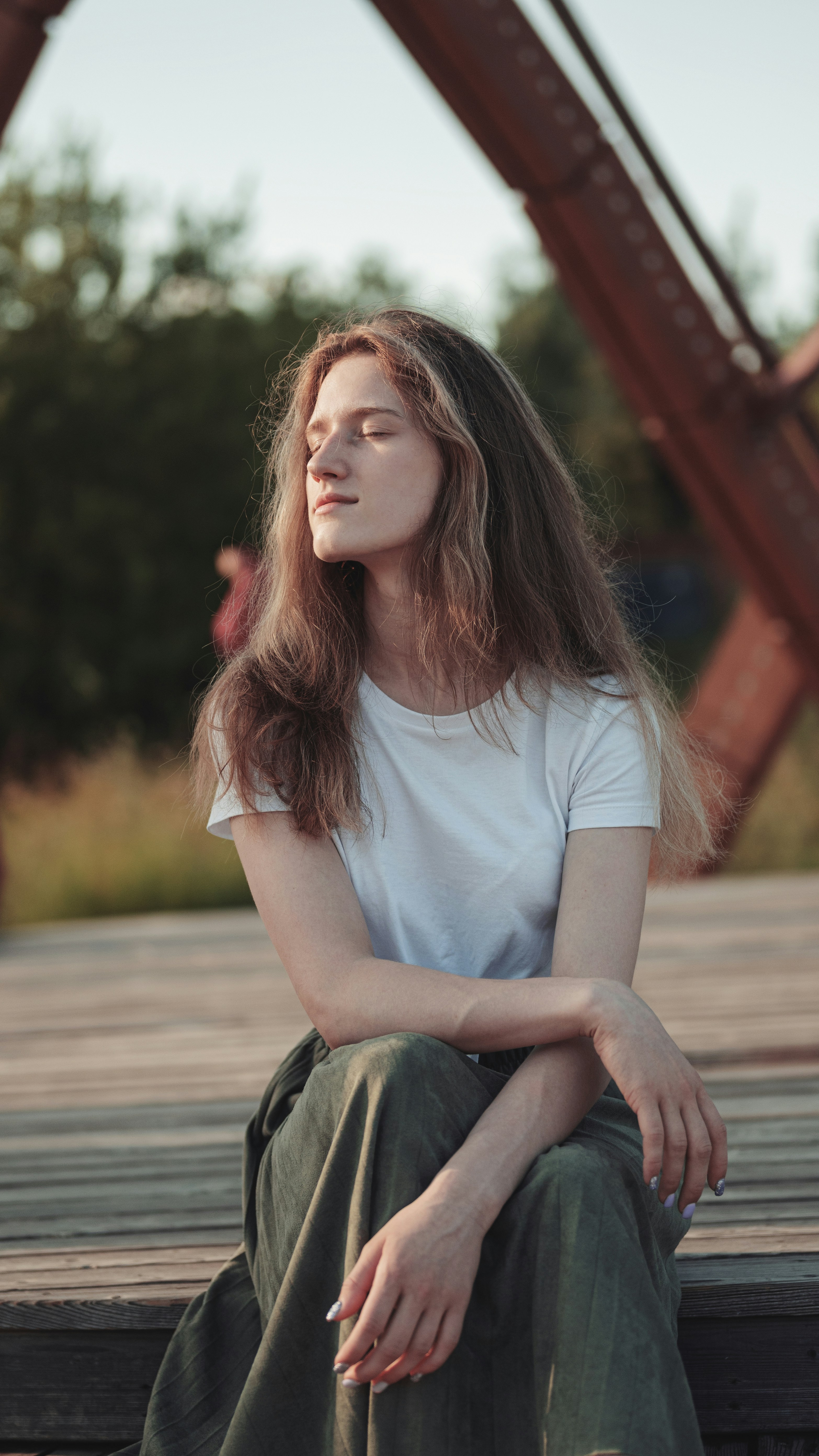 A young woman with long hair sits peacefully on a wooden platform, basking in the sunlight, surrounded by greenery and industrial structures.
