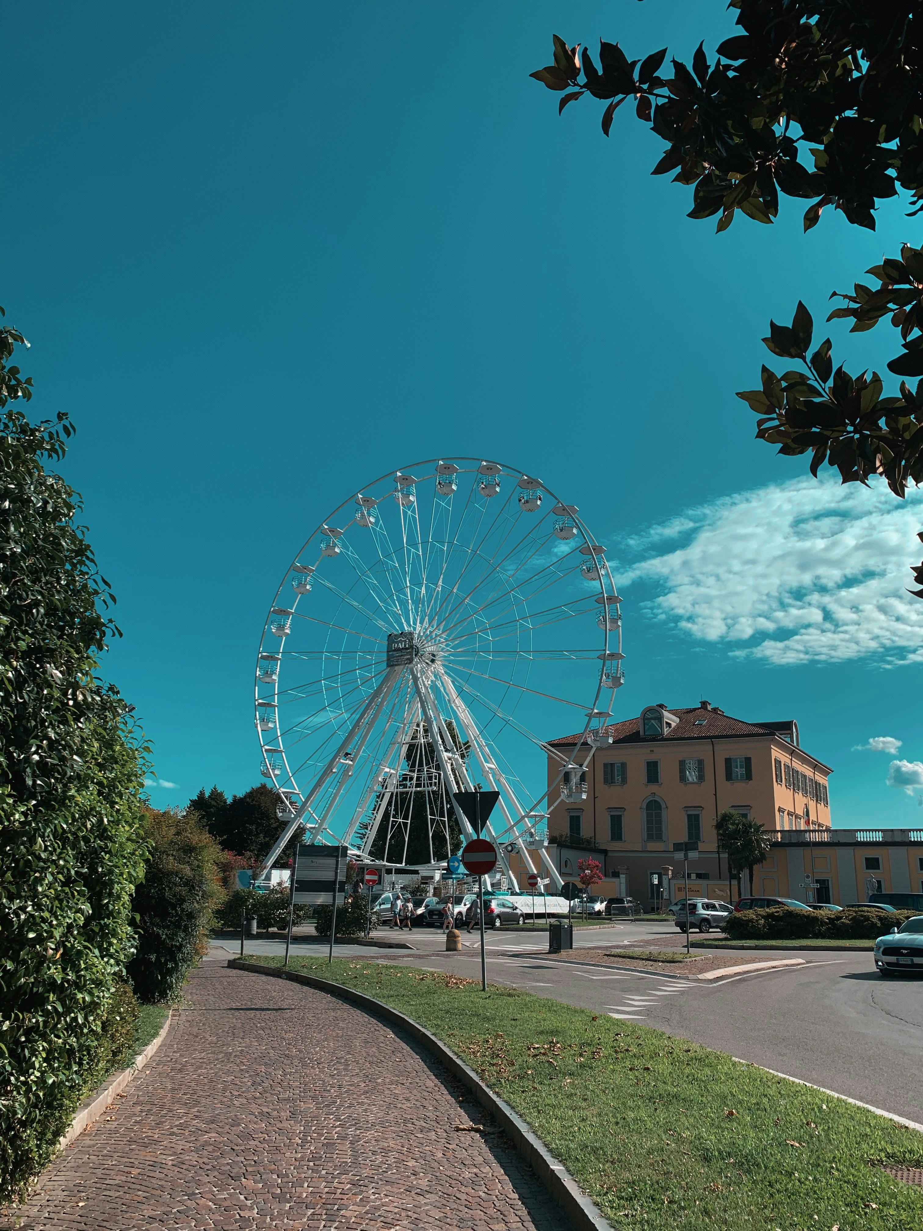 A vibrant Ferris wheel stands tall against a clear blue sky, surrounded by lush greenery and charming architecture. The scene captures the essence of a lively outdoor fairground.