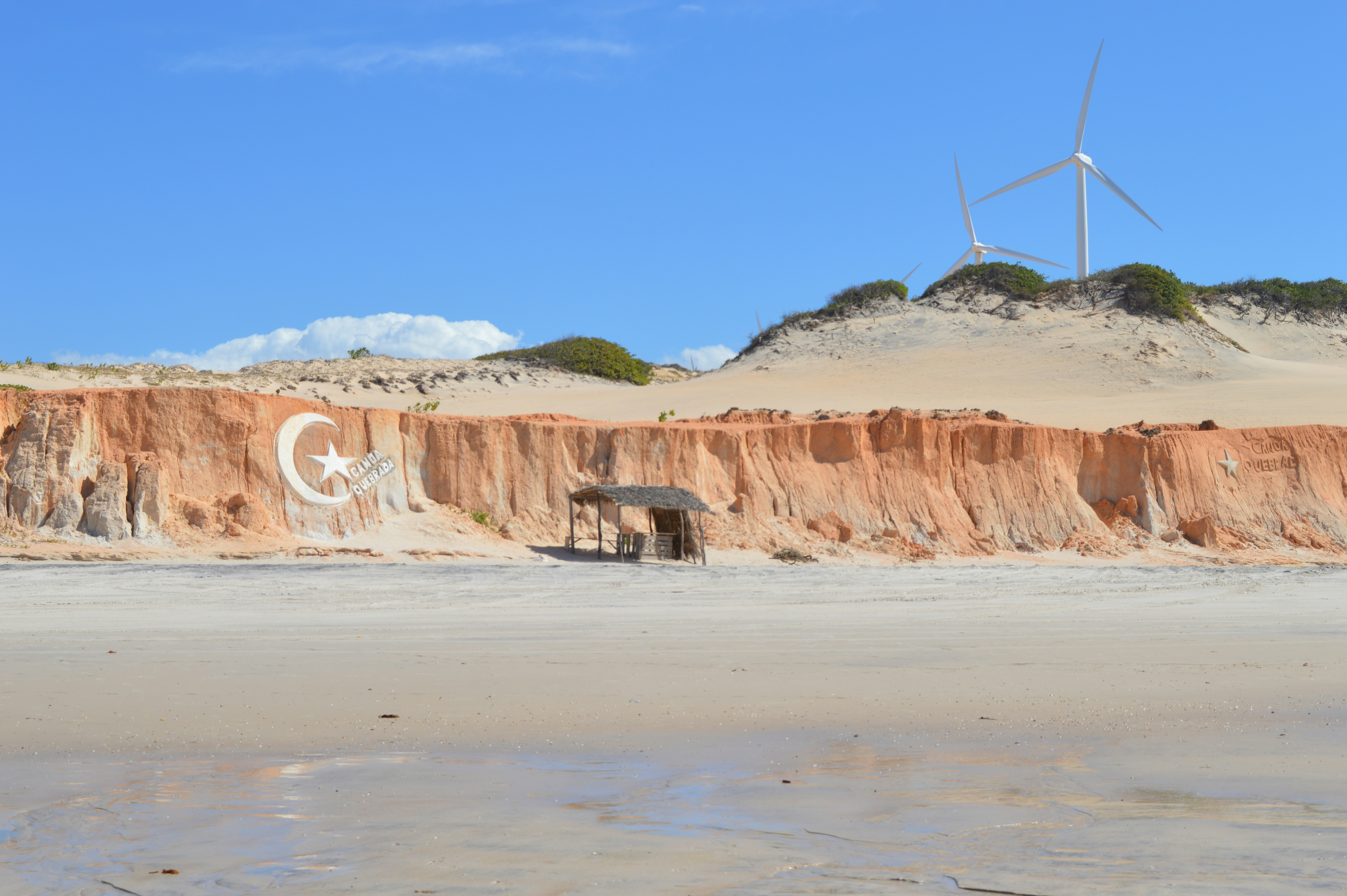Sandy cliffs with a wind turbine above and a calm beach in the foreground under a clear blue sky.