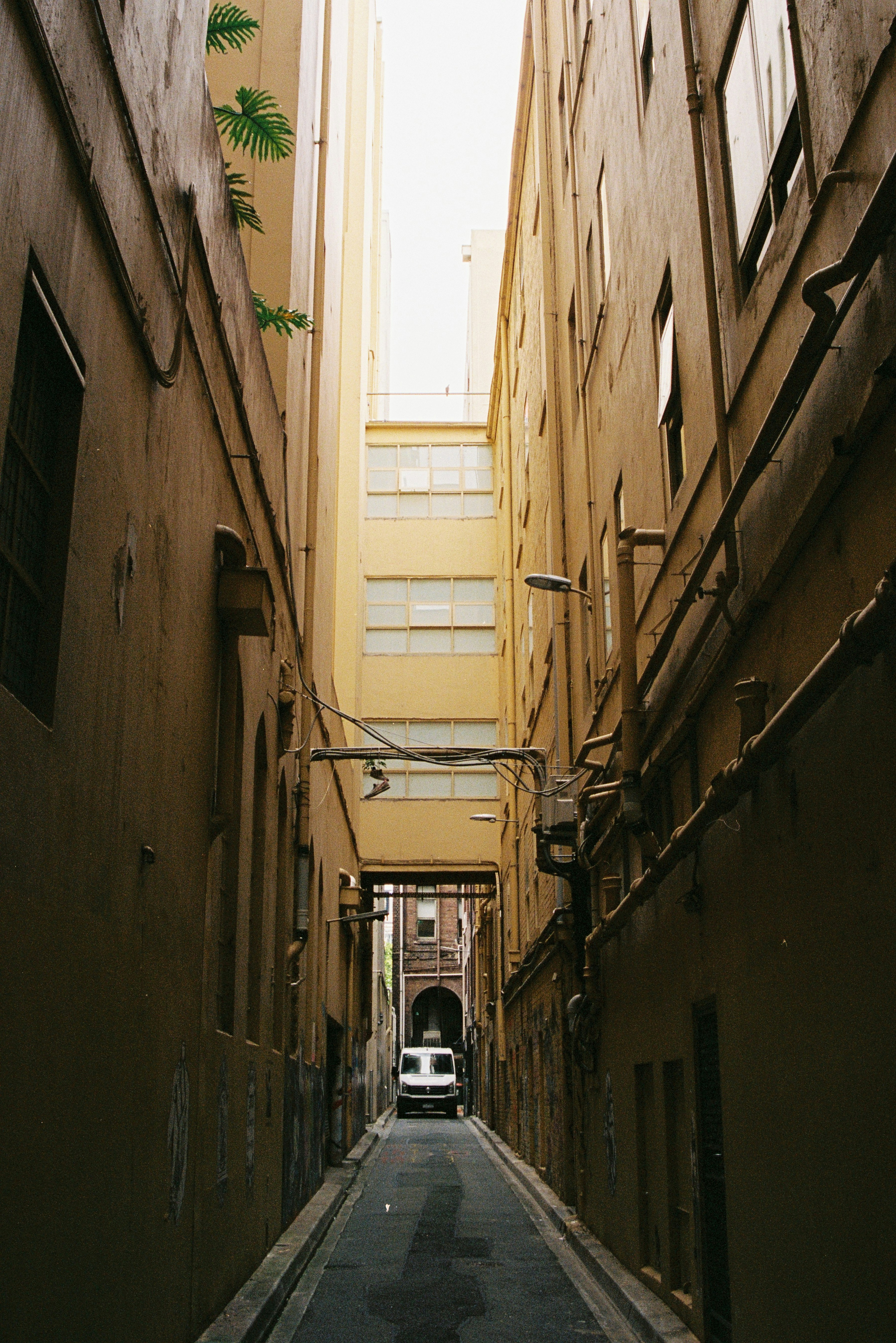 Narrow alleyway flanked by tall yellow buildings, leading to a van parked at the far end. The scene captures the essence of urban exploration.