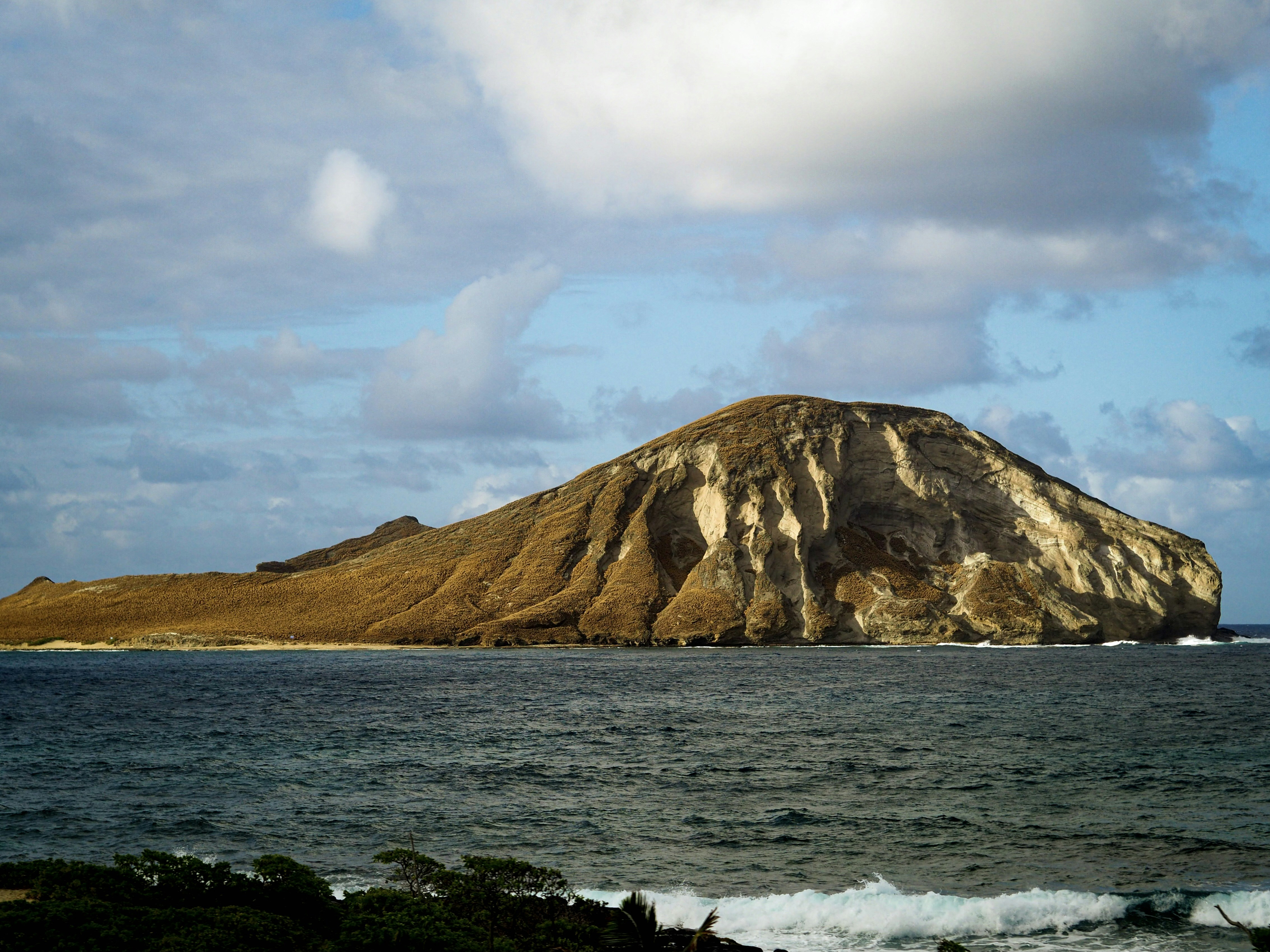 A rugged island rises from the ocean, showcasing its textured cliffs and golden slopes under a dynamic sky. Waves gently lap at its base.