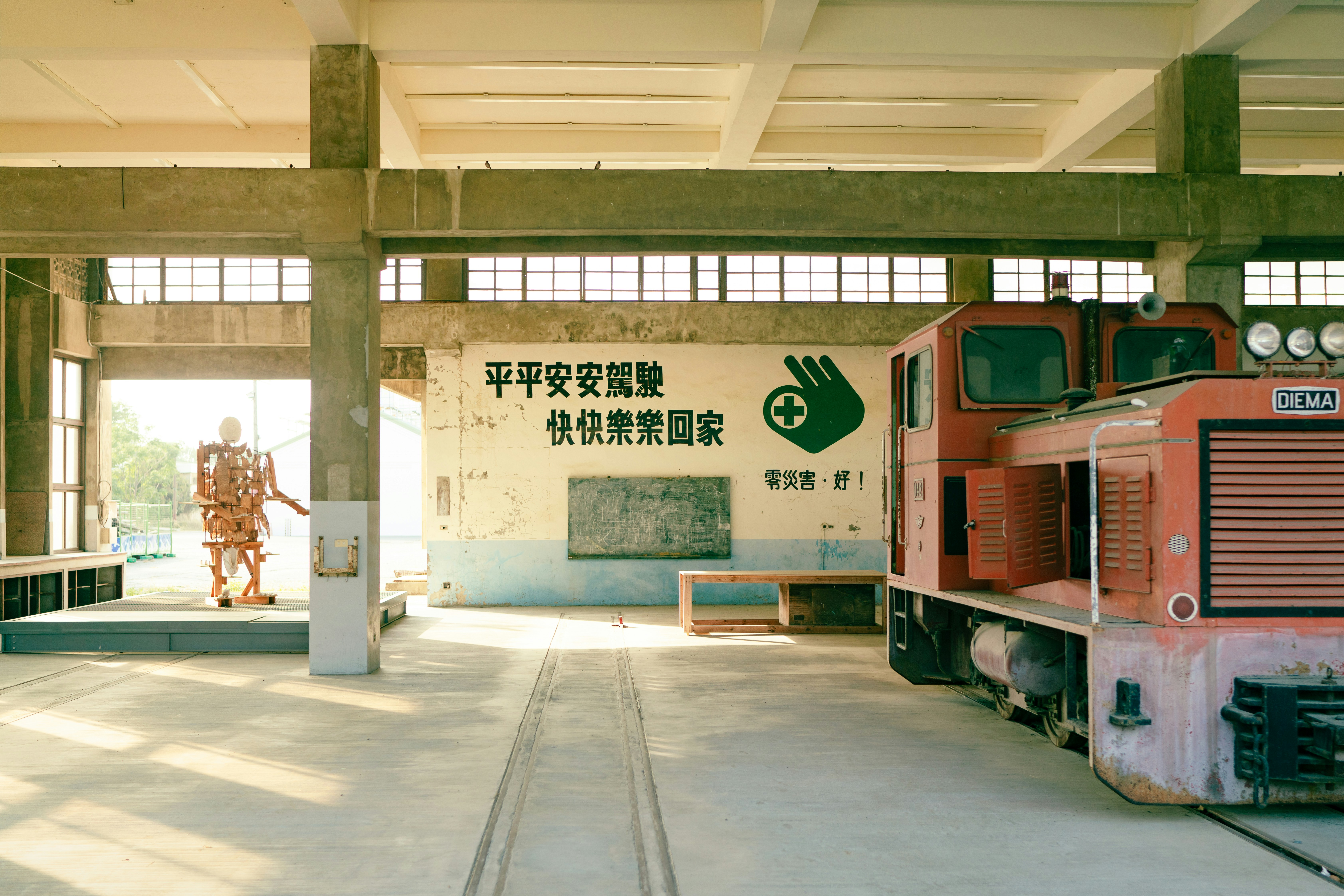 An abandoned industrial space featuring a vintage train and a humanoid sculpture, illuminated by soft natural light. The walls bear faded signage, hinting at a once-bustling environment.