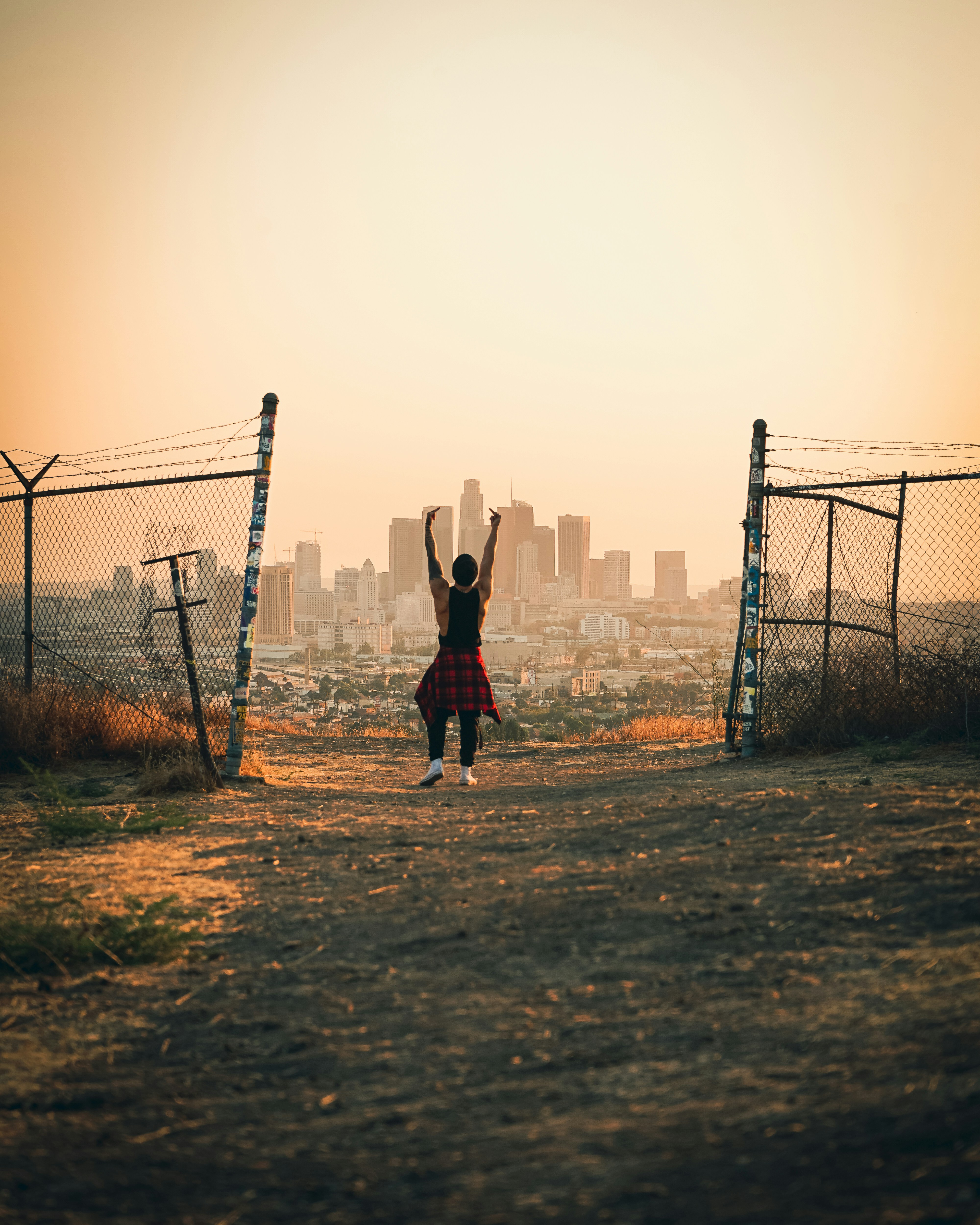 Silhouette of a person celebrating with arms raised in front of a sprawling city skyline at sunset, framed by an open gate.