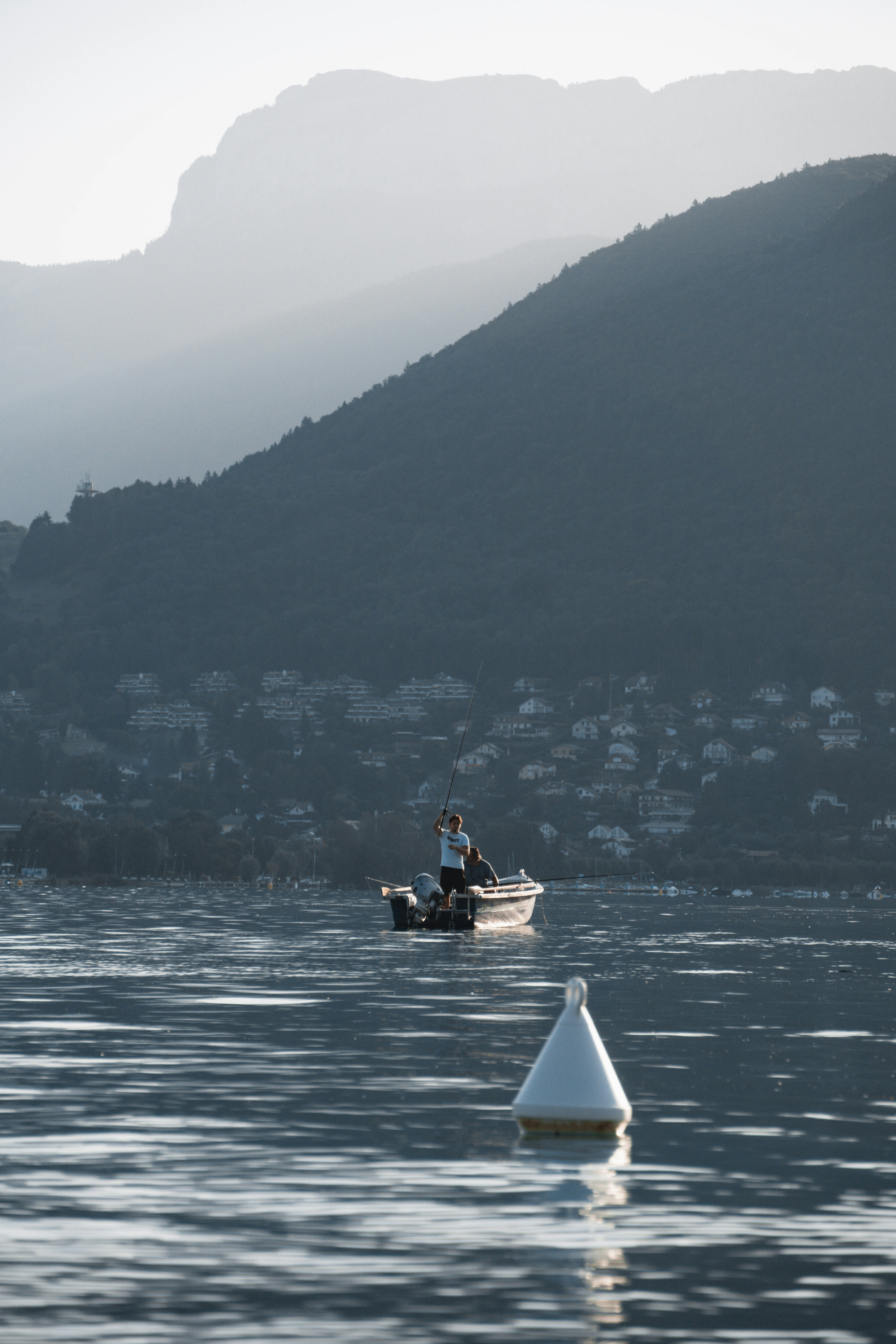 A lone fisherman casts his line from a small boat, surrounded by tranquil waters and distant mountains. A buoy floats nearby.