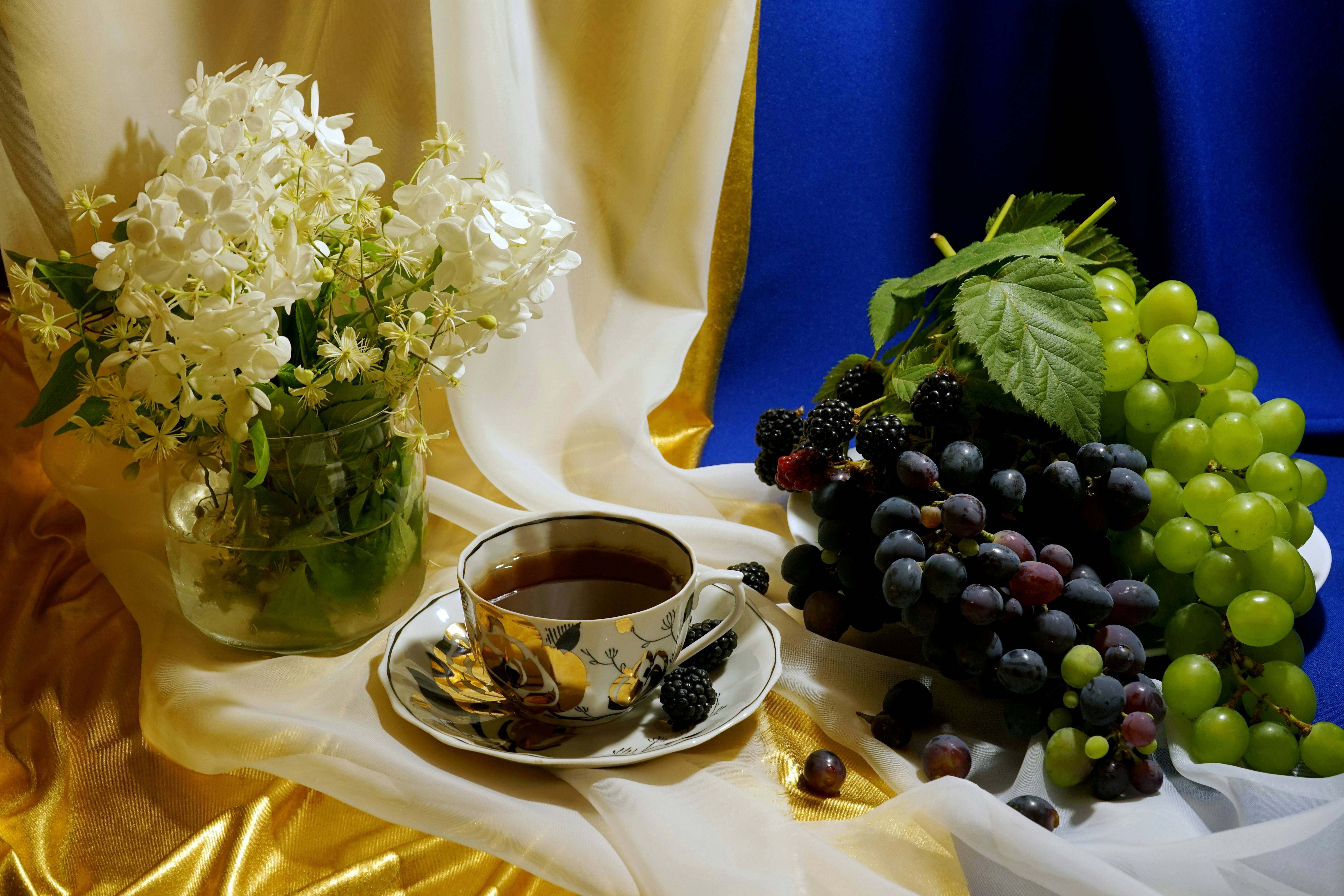 A delicate arrangement featuring a teacup beside a lush assortment of grapes and a vase of white flowers, set against rich fabric drapes.