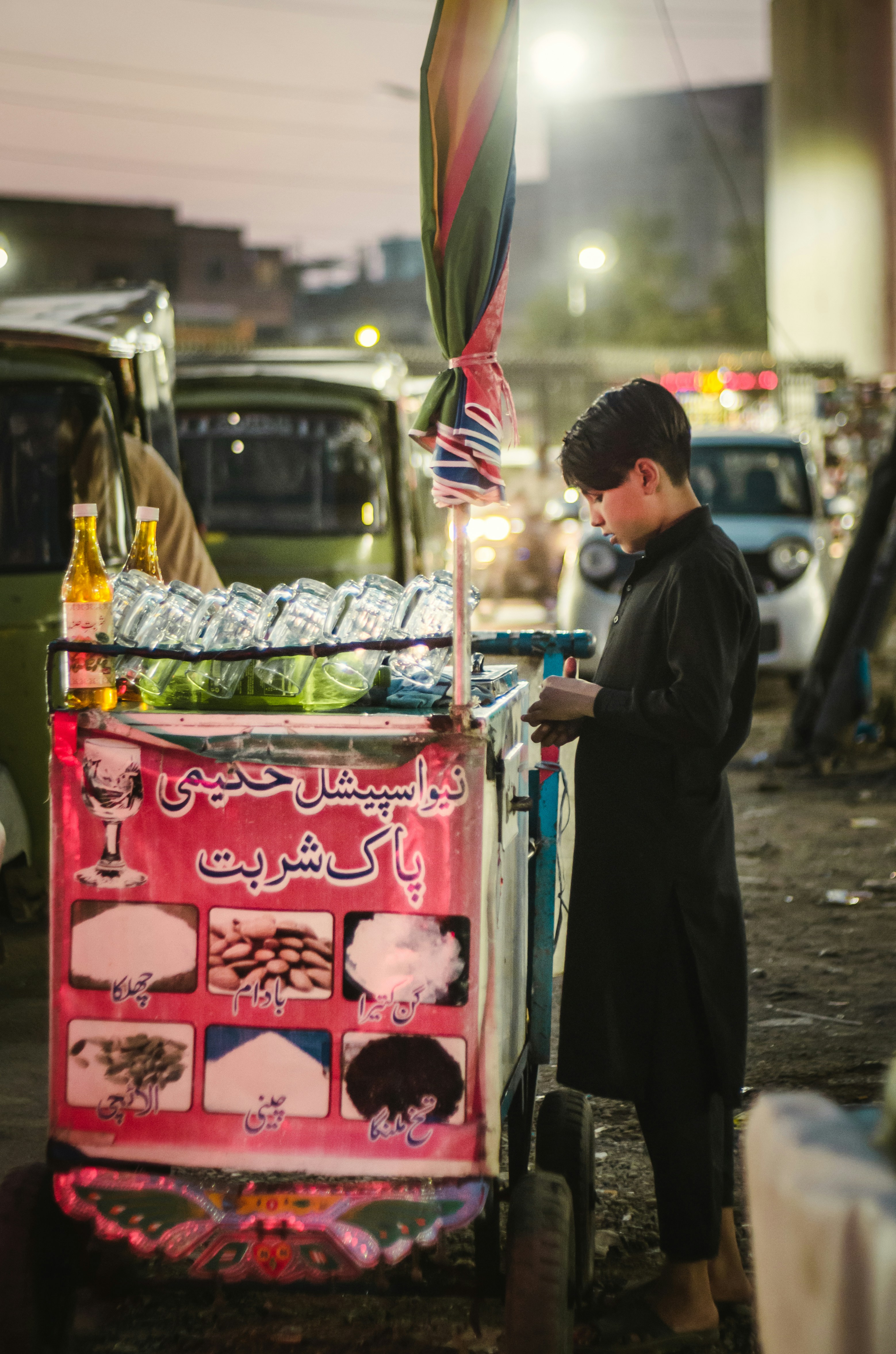 A street vendor in a dark traditional outfit stands beside a pink cart topped with water bottles, illuminated by a dim market glow.