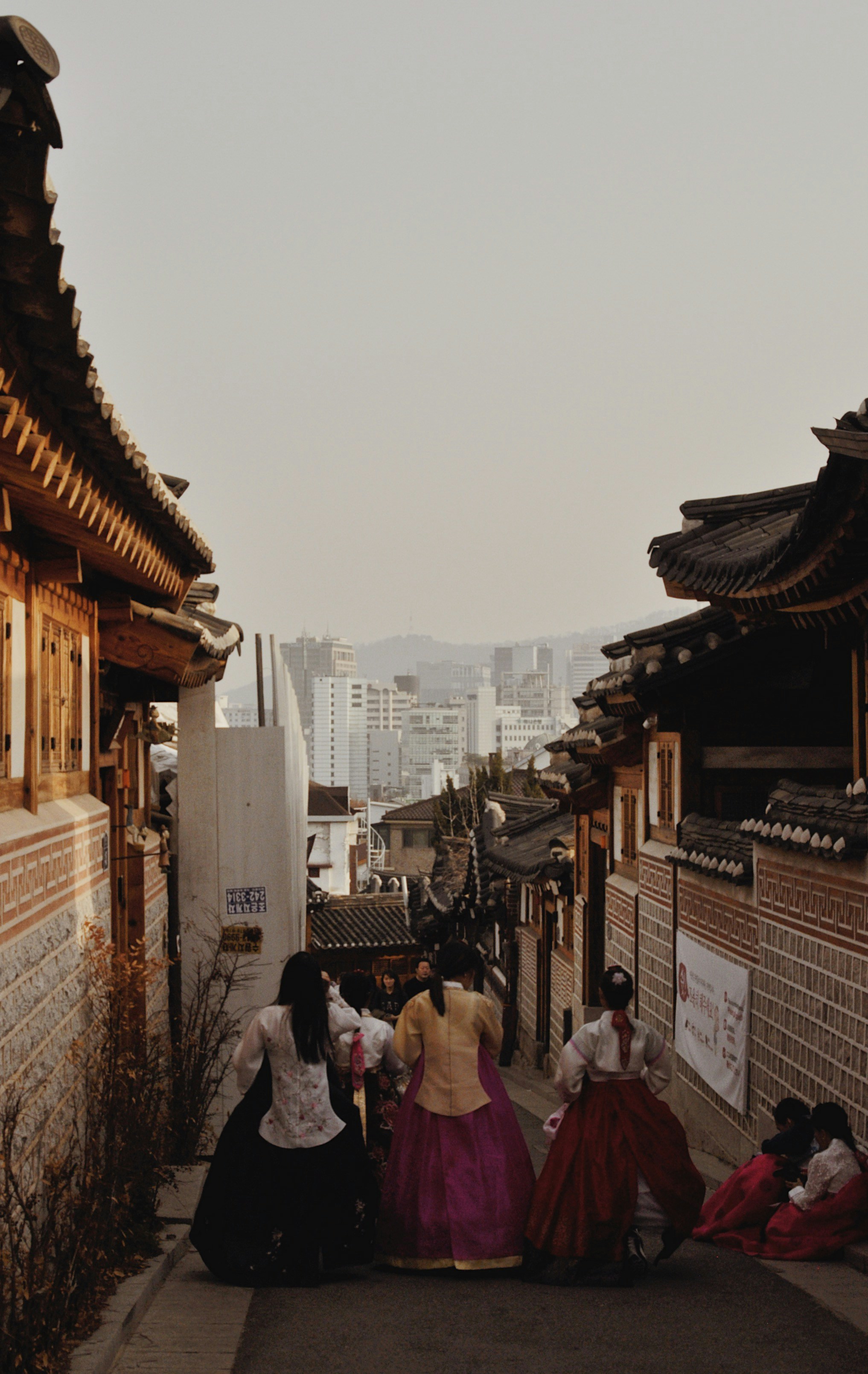 Group of individuals in traditional hanbok attire walking down a historic street, framed by traditional Korean architecture and a modern city skyline in the background.