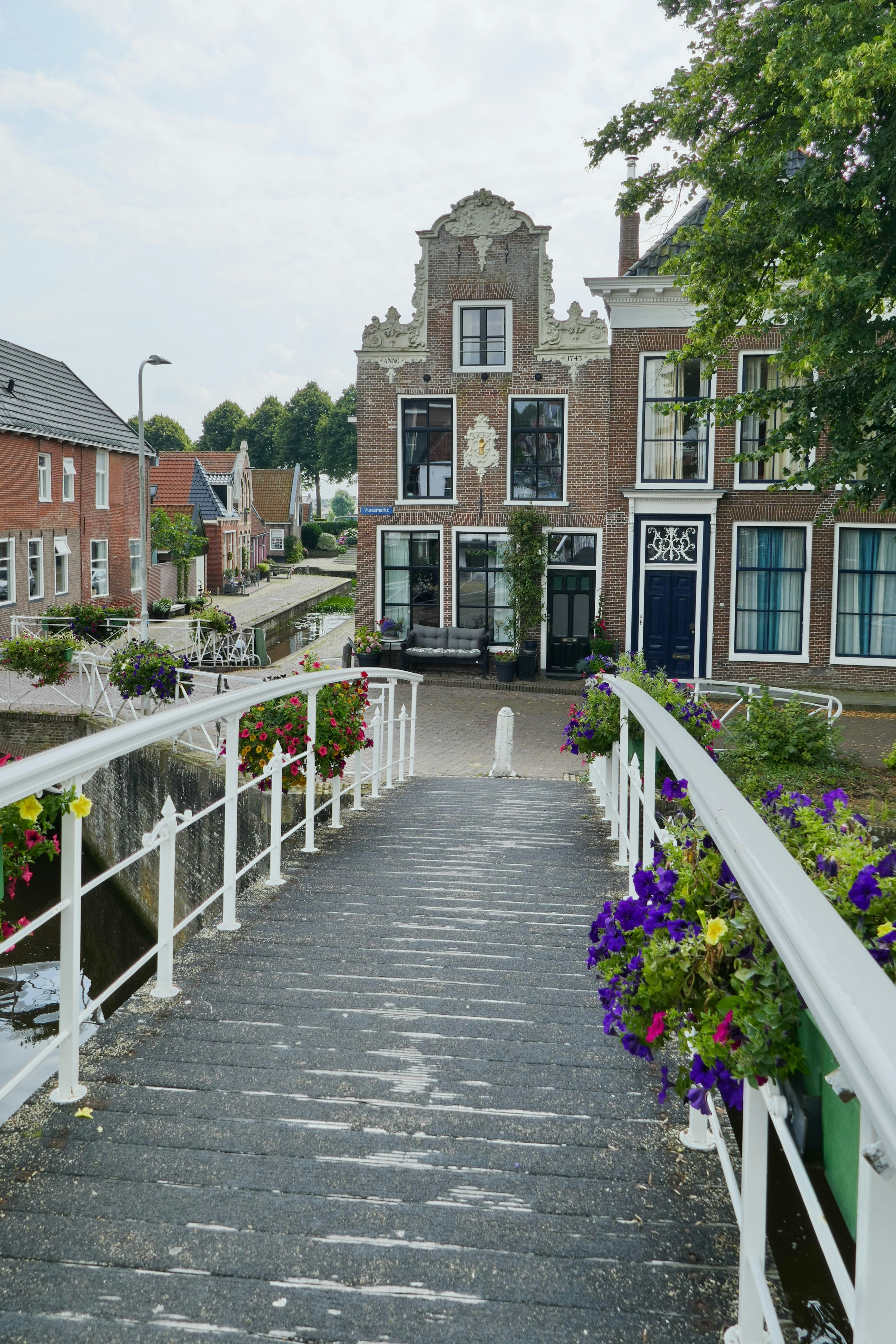 A wooden bridge adorned with vibrant flowers leads to a picturesque canal-side house in a serene village setting.