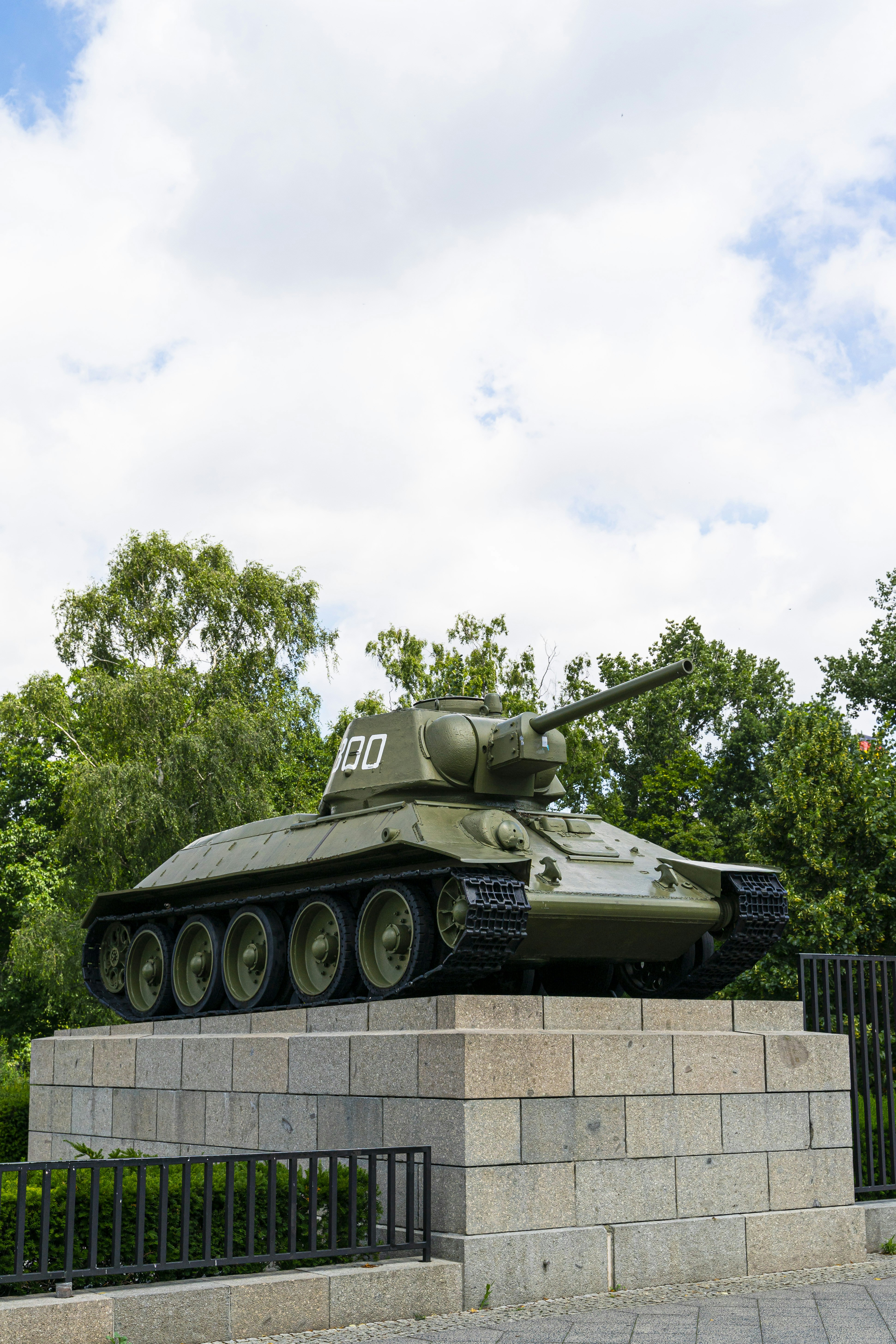 Soviet T-34 tank monument on a stone pedestal, surrounded by lush greenery and a clear sky.