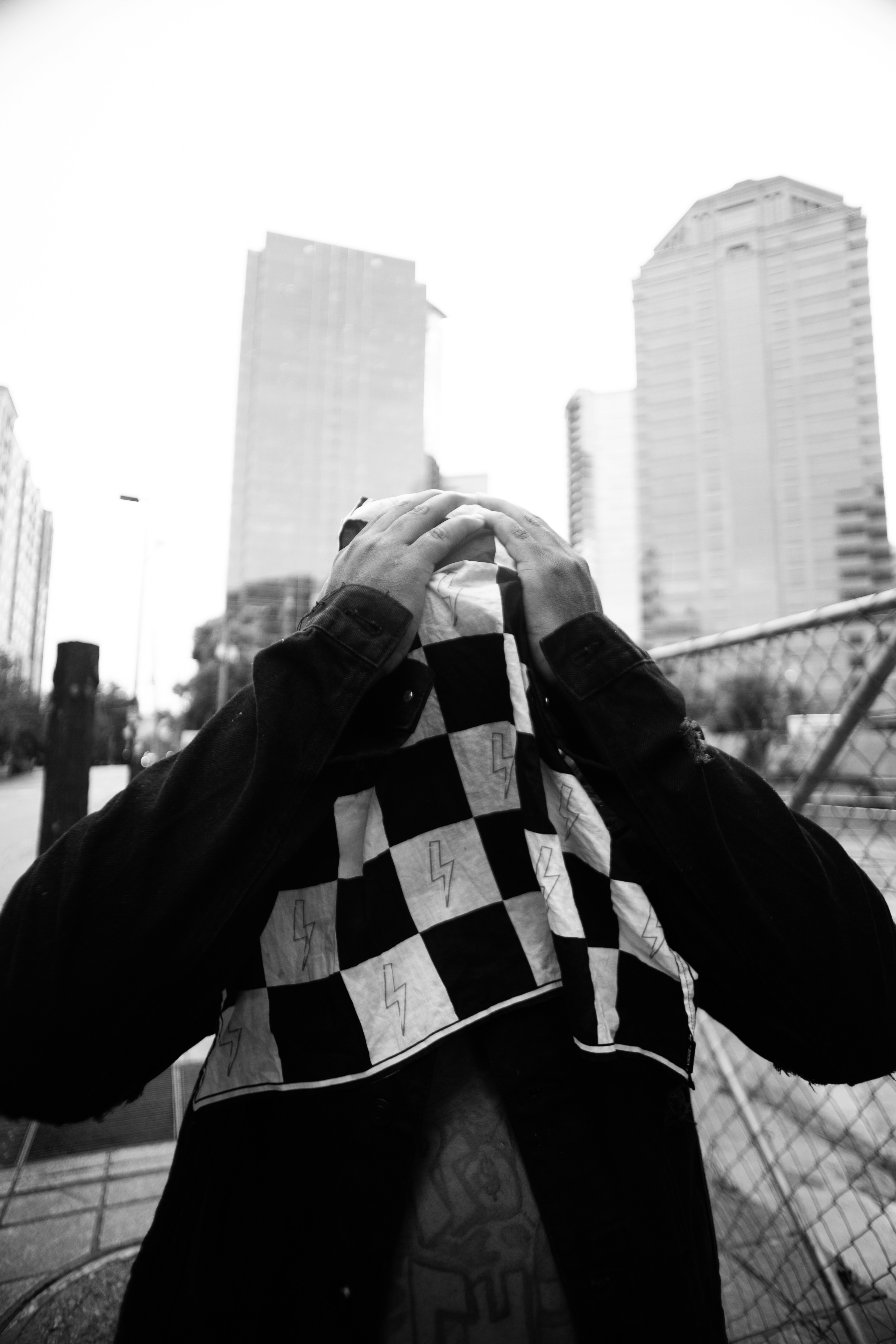 Individual obscured by a checkered cloth stands against a backdrop of towering city buildings, evoking a sense of isolation in an urban landscape.