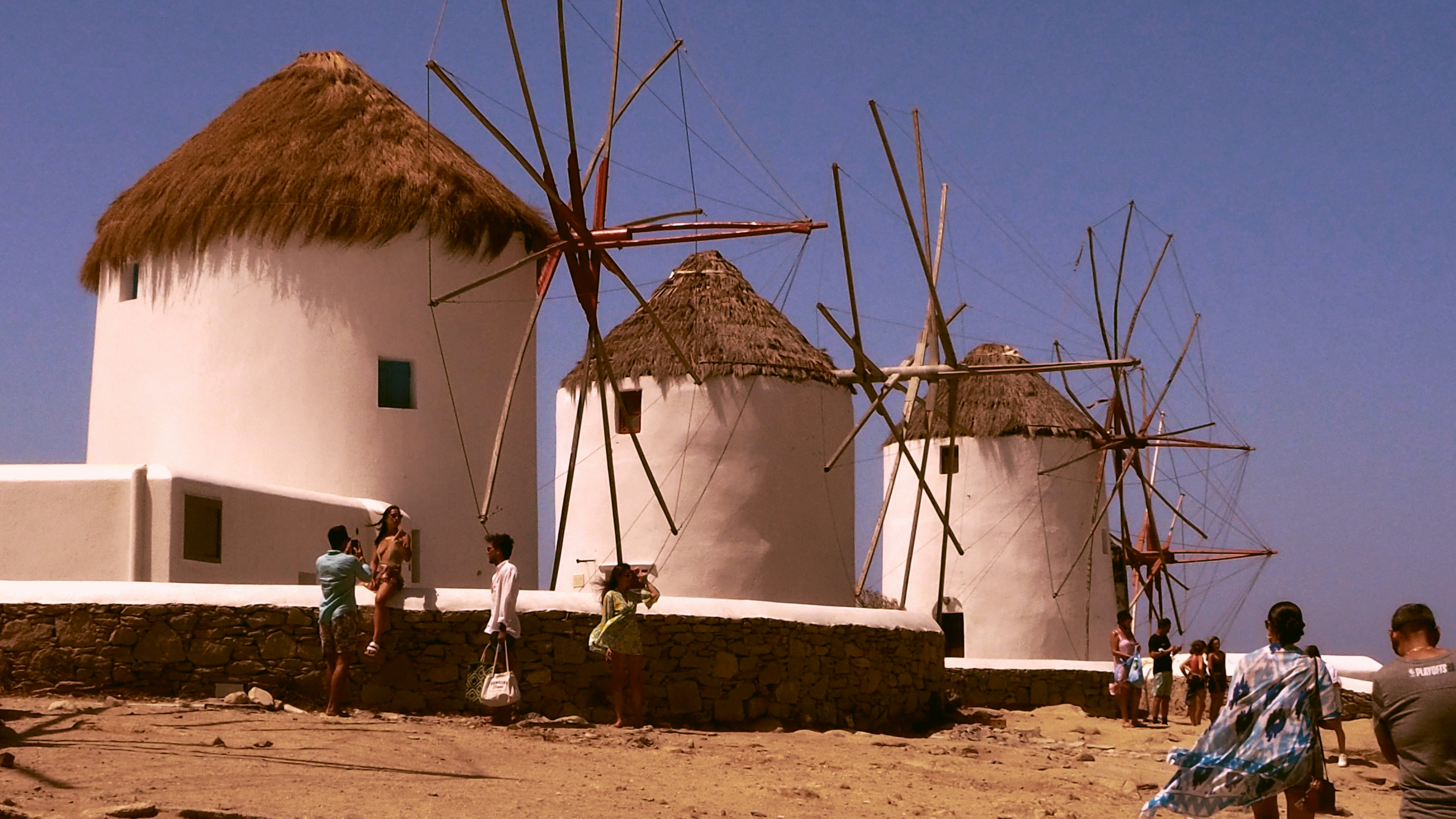 Three traditional windmills with thatched roofs stand against a clear blue sky, surrounded by visitors enjoying the scenic coastal atmosphere.